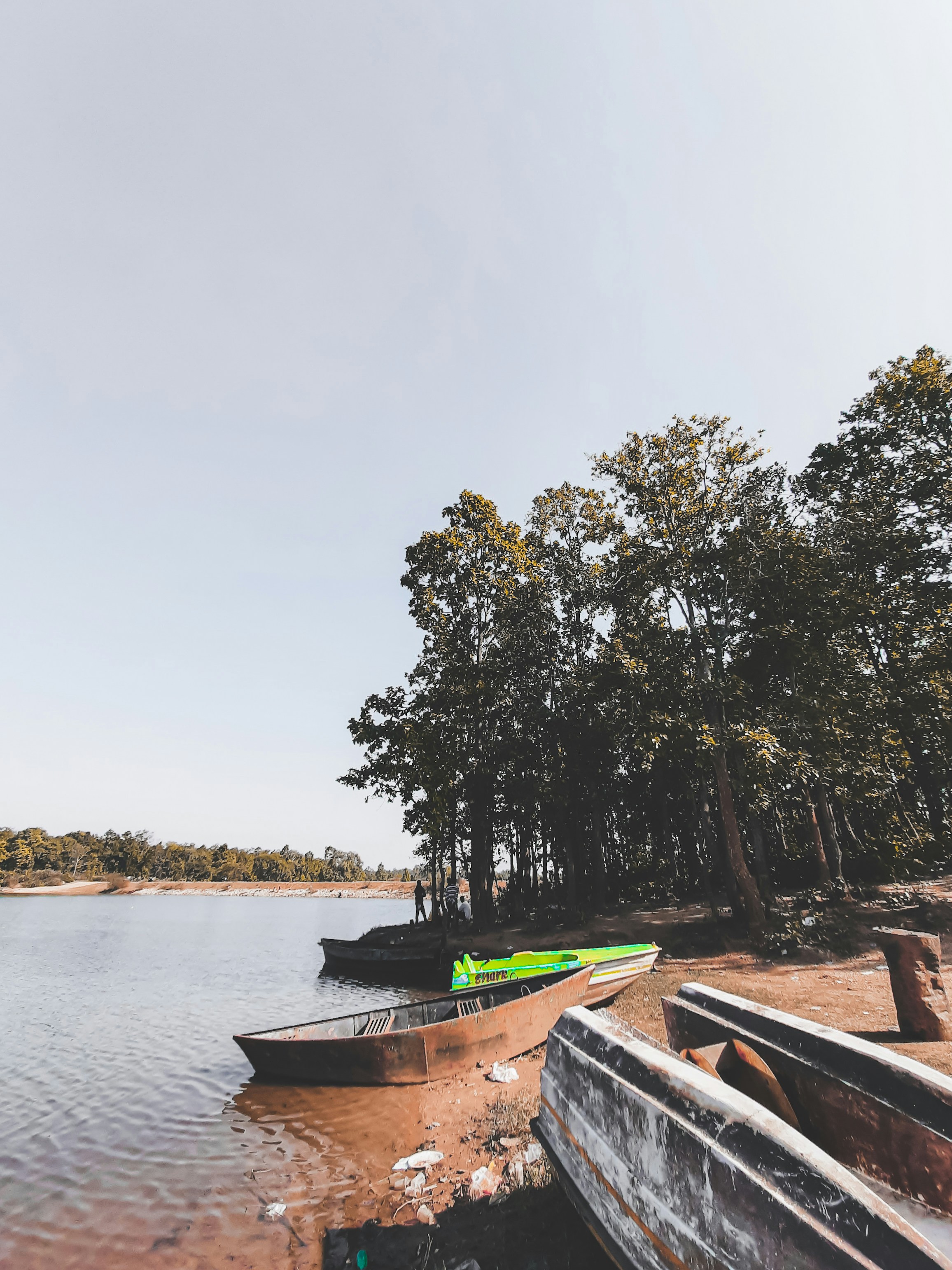 Two boats resting on the shore beside a calm river, framed by lush green trees under a clear sky.