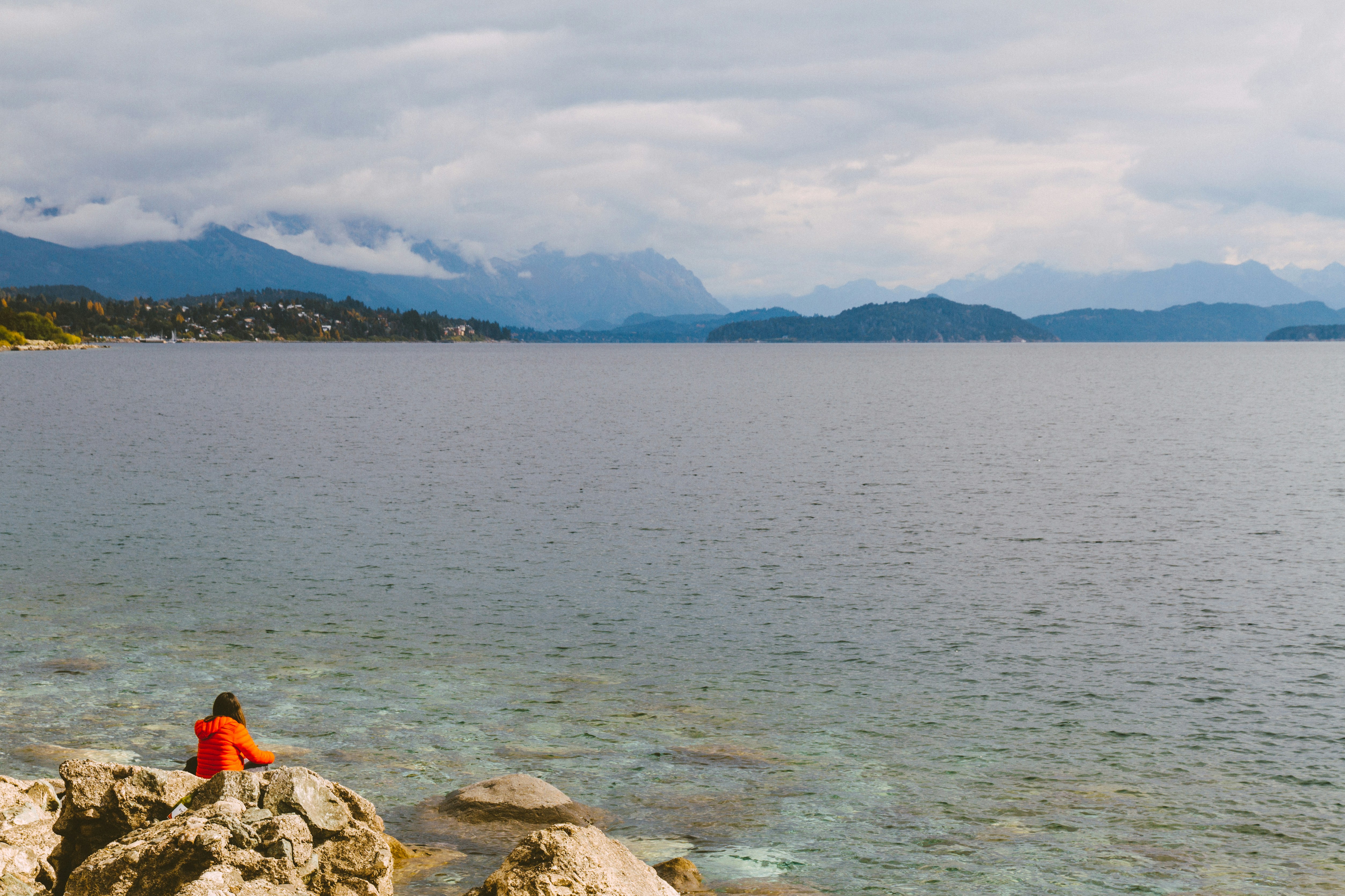 Person in red jacket sitting on rocky shoreline overlooking a calm lake with distant mist-covered mountains.
