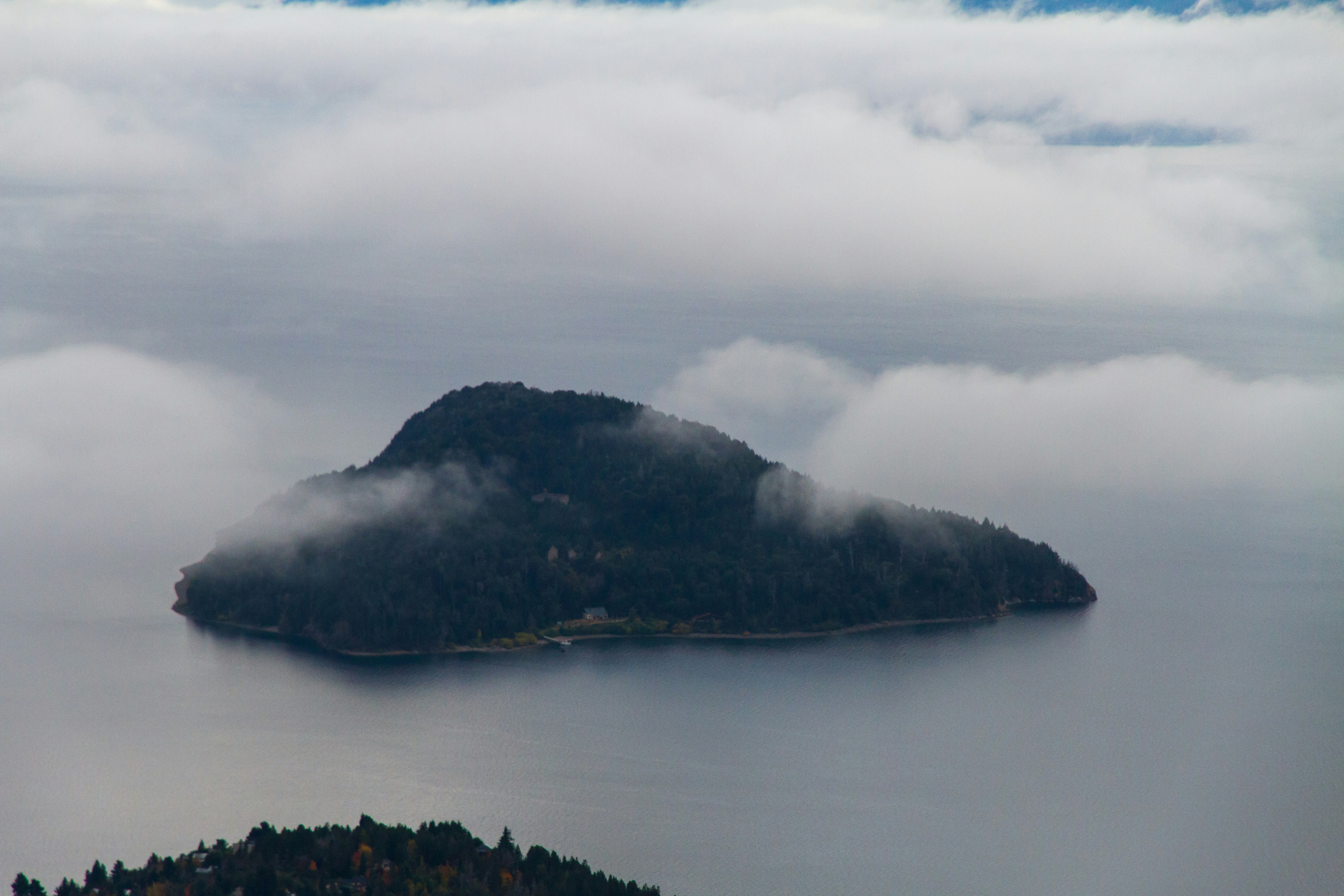 A solitary island emerges from a tranquil lake, shrouded in soft clouds and mist, creating a serene atmosphere.