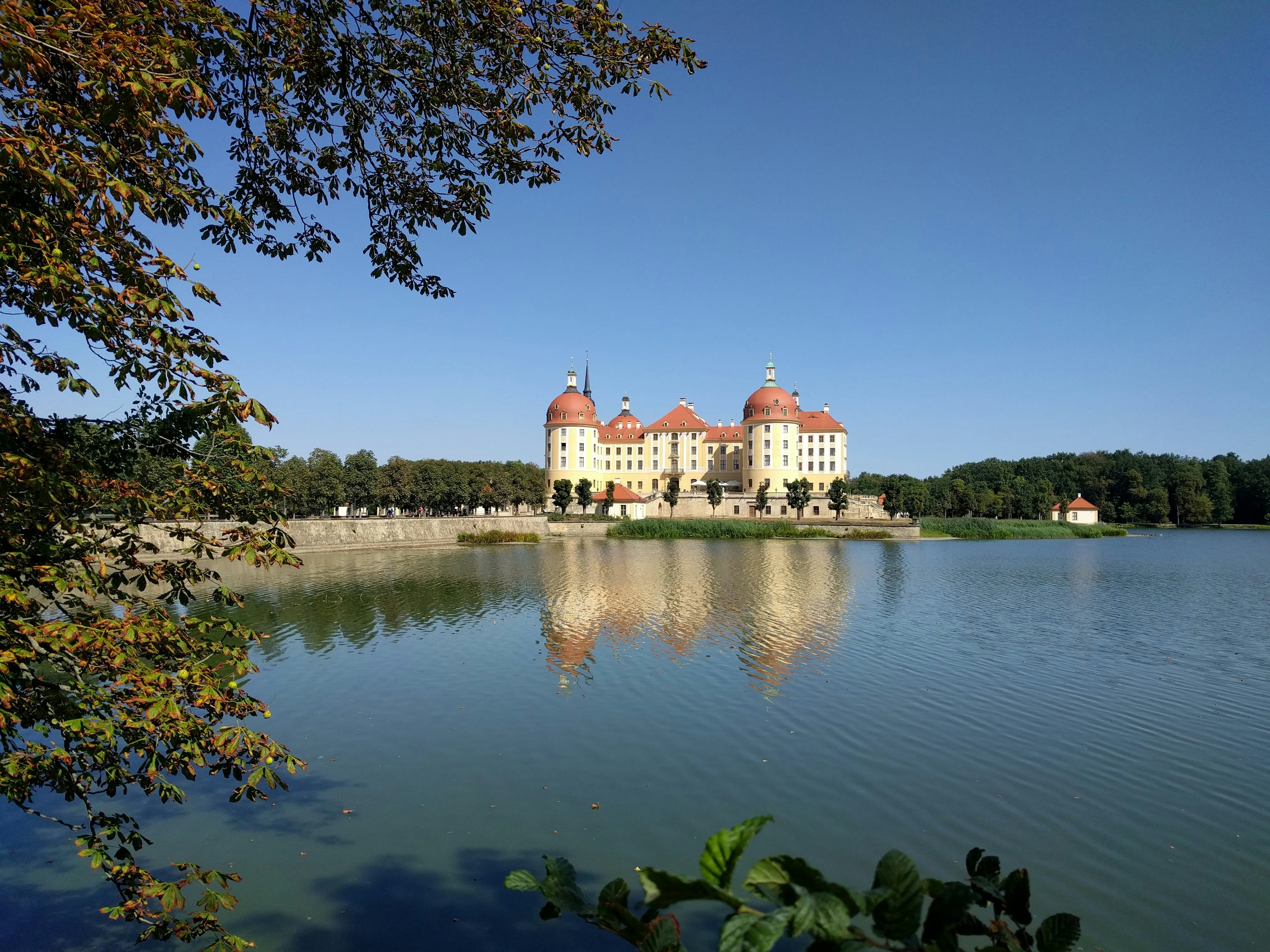 Historic castle with red domes reflected in a serene lake, surrounded by lush greenery and clear blue skies.
