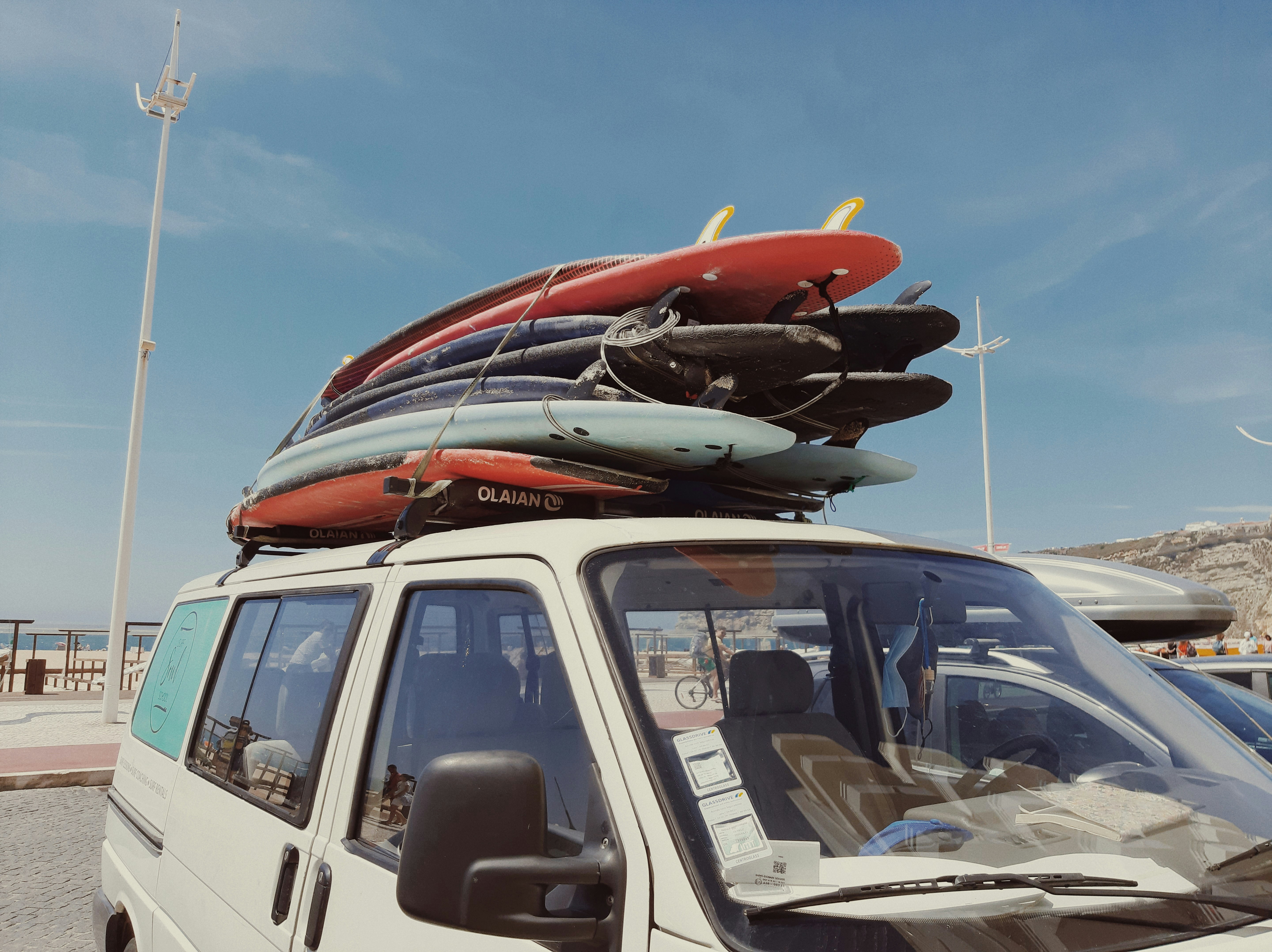 A white van parked in a seaside lot with a roof rack stacked high with colorful surfboards under a clear blue sky.