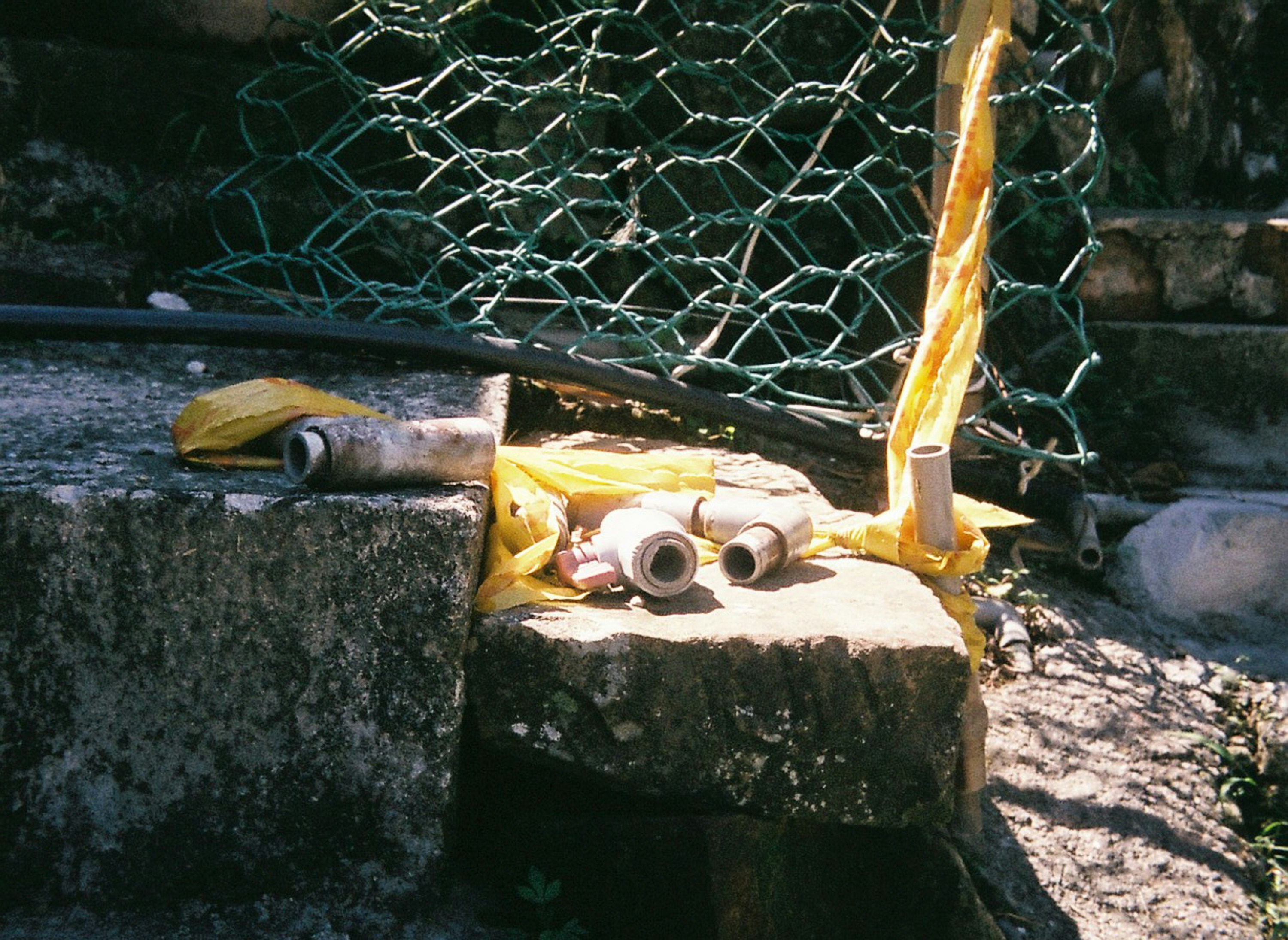 A collection of discarded pipes and yellow plastic scattered on a stone surface, surrounded by a green netting and natural elements. 