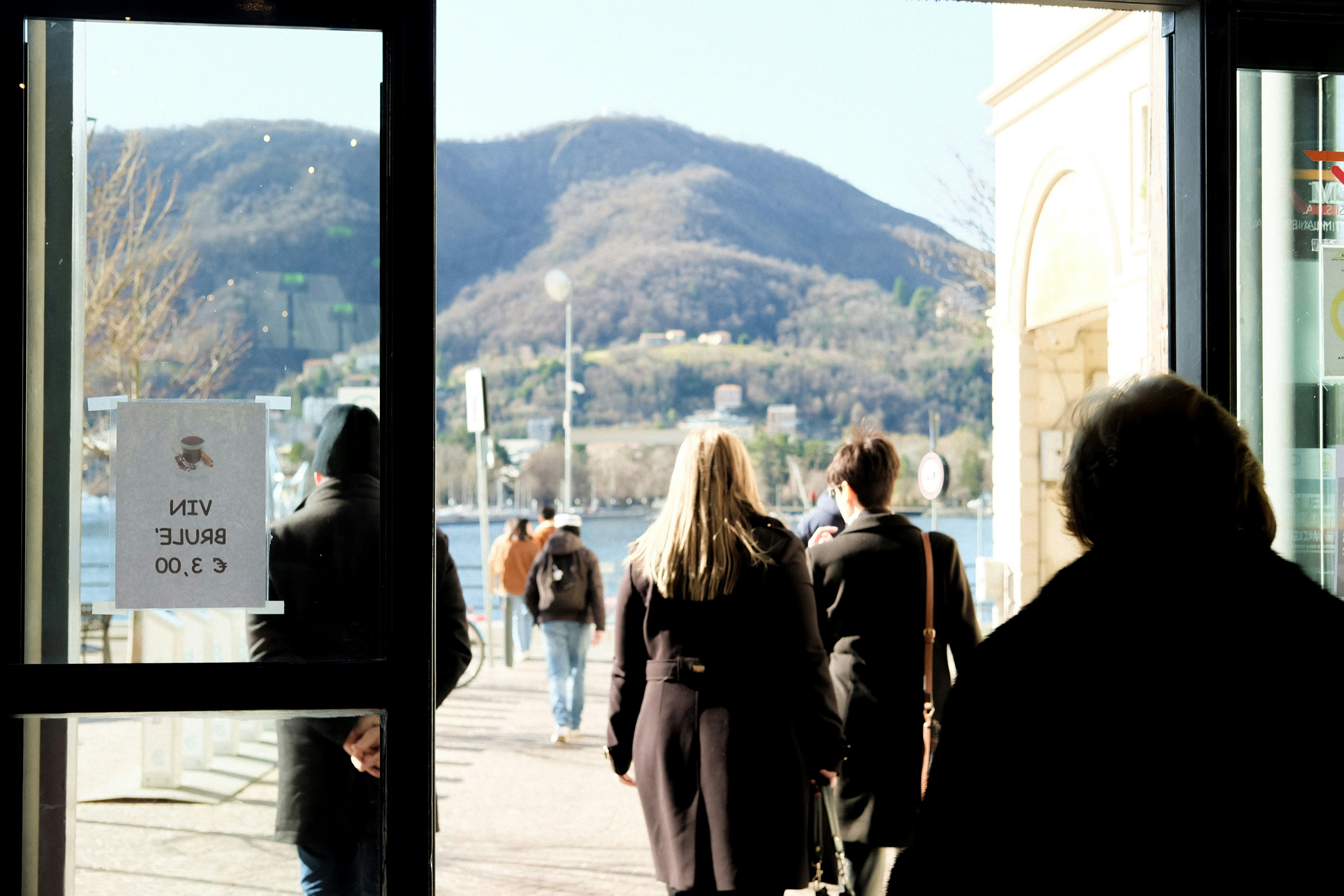 People walking towards a sunlit mountain view through a glass doorway.