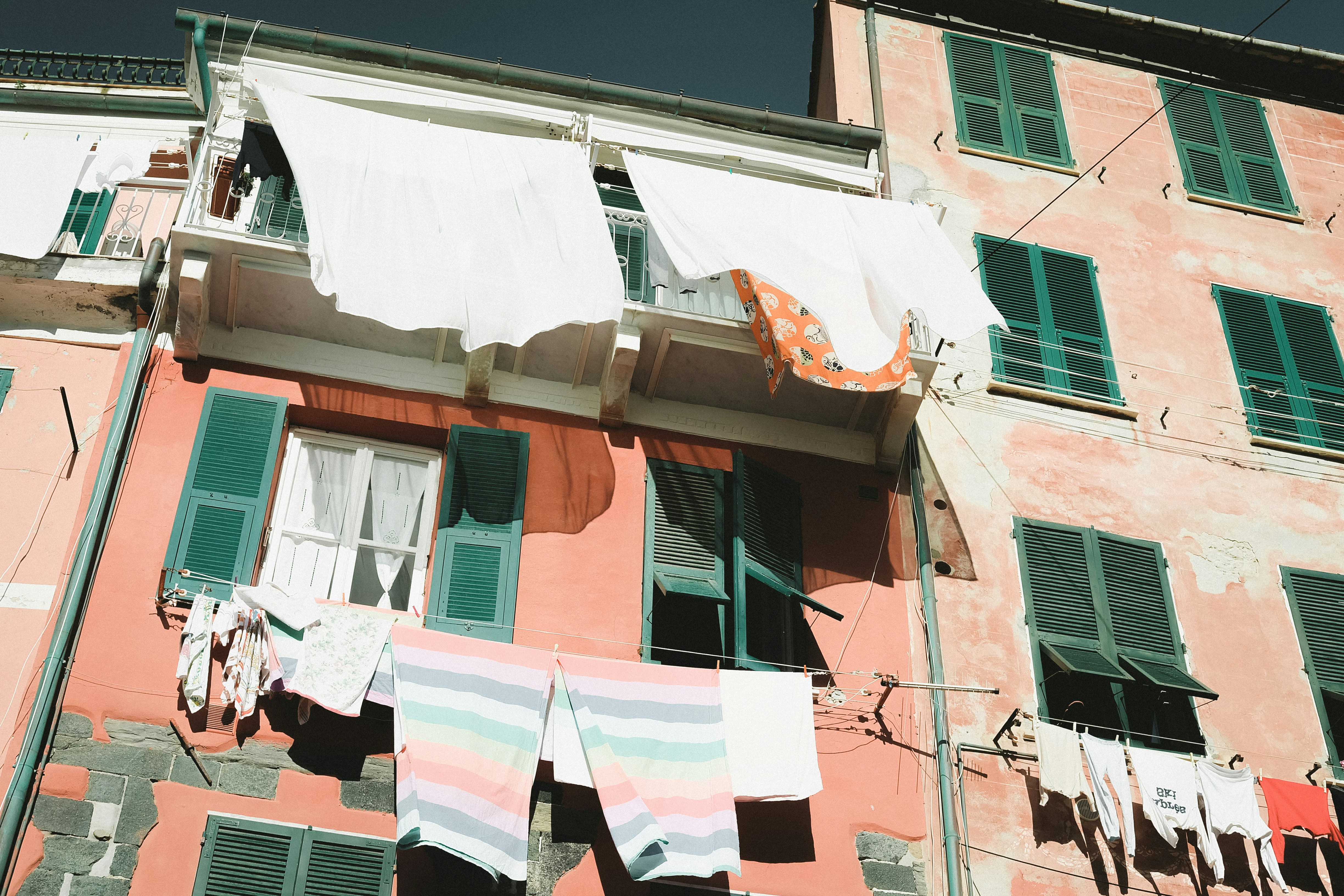 Colorful laundry hangs against a sunlit pink facade with green shutters.