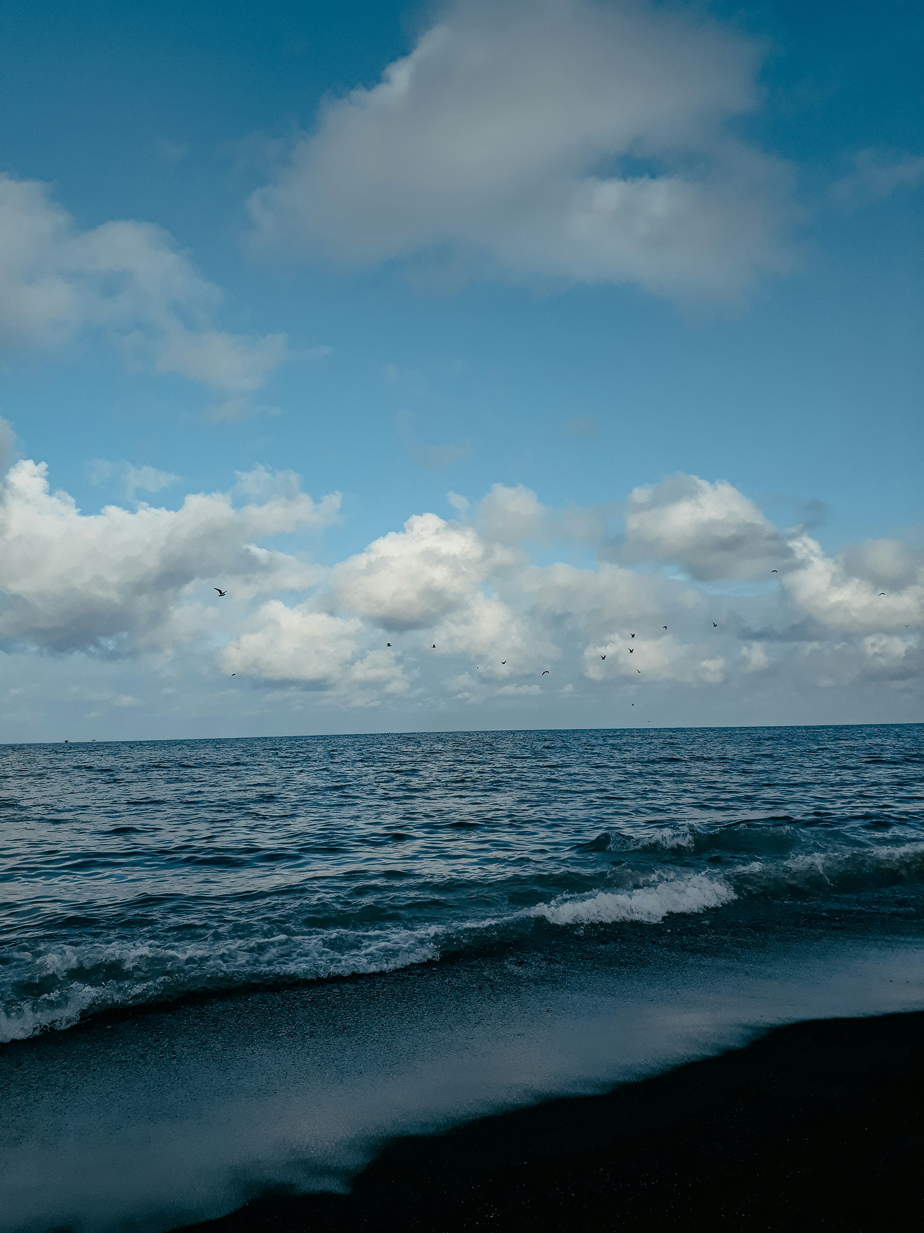 Calm sea stretches to a pale horizon beneath a blue sky dotted with clouds. Dark sand in the foreground anchors the composition.