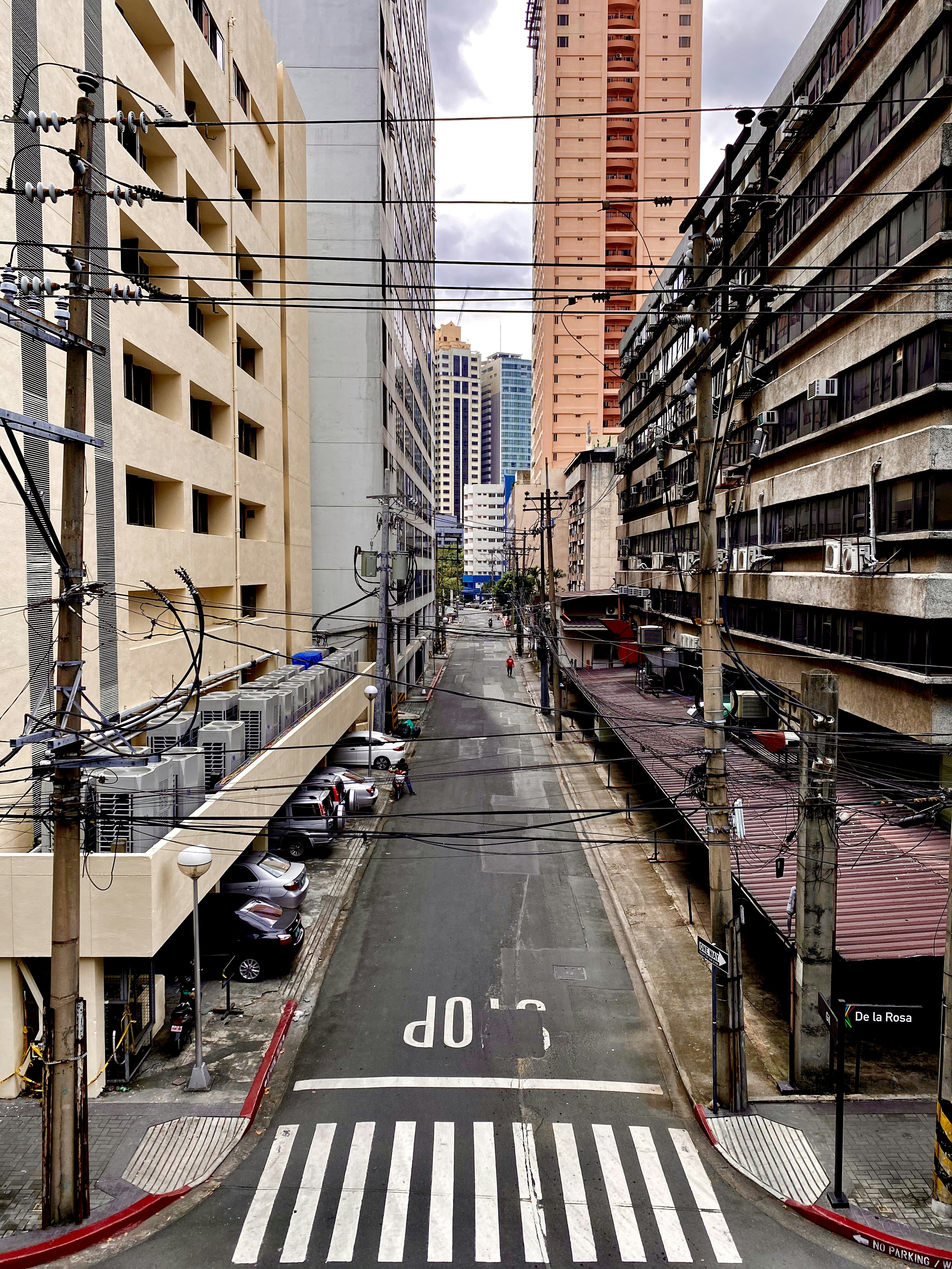 A city street lined with towering buildings and overhead power lines, showcasing a blend of modern architecture and urban life. The scene is punctuated by a stop sign and crosswalk.