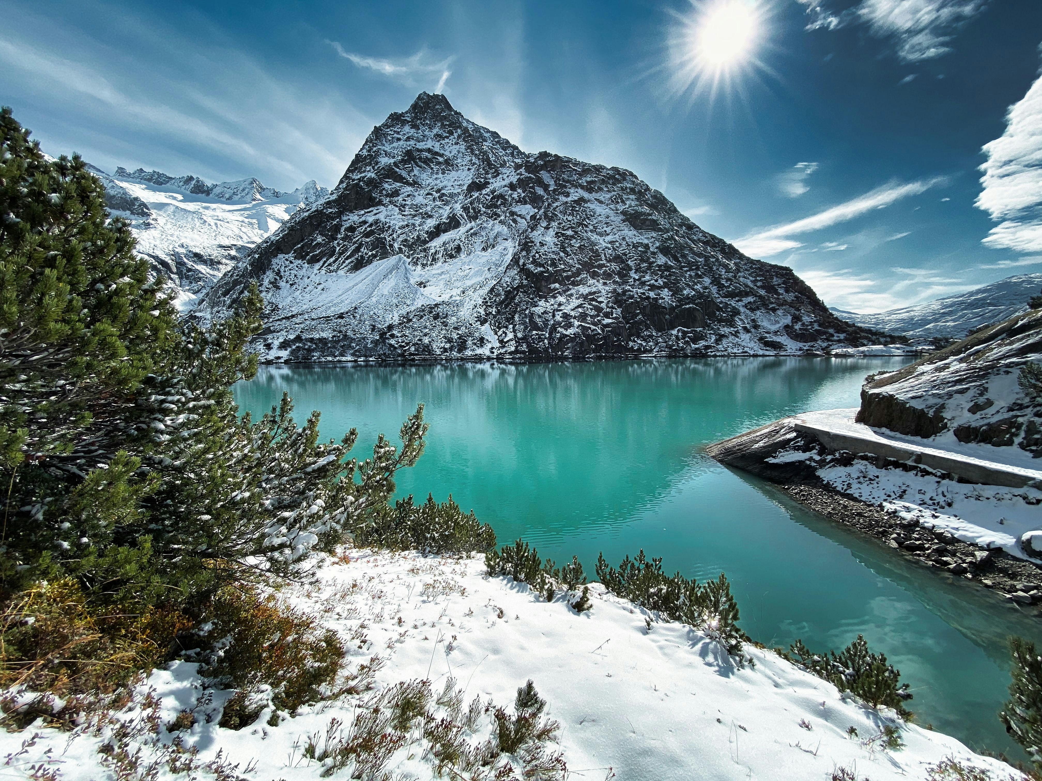 Snow-covered mountains rise majestically above a turquoise lake, reflecting the vibrant sky. Pine trees frame the serene landscape.