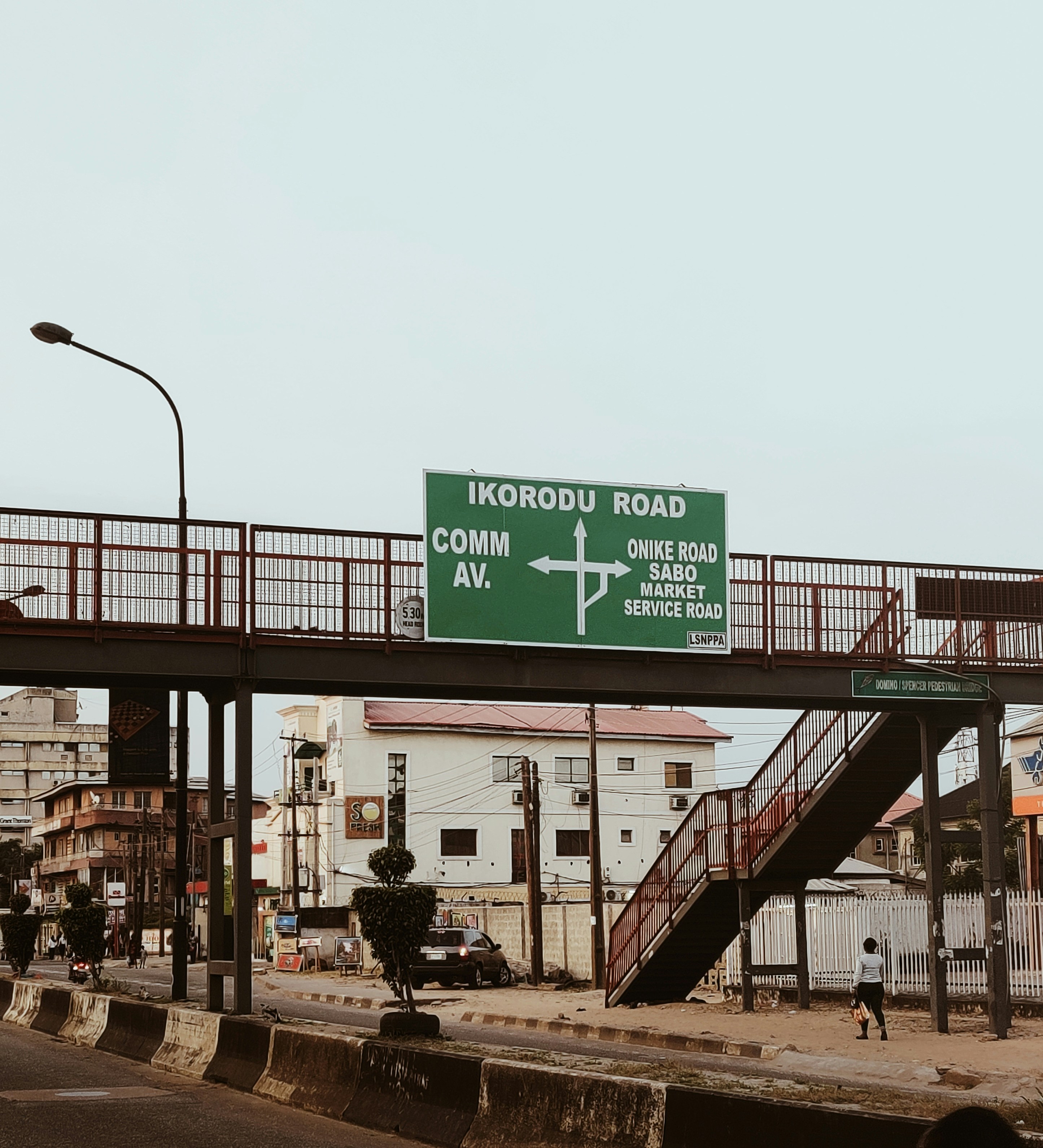 Signpost indicating directions on Ikorodu Road with pedestrian overpass in view. Urban scene showcasing local architecture and traffic.