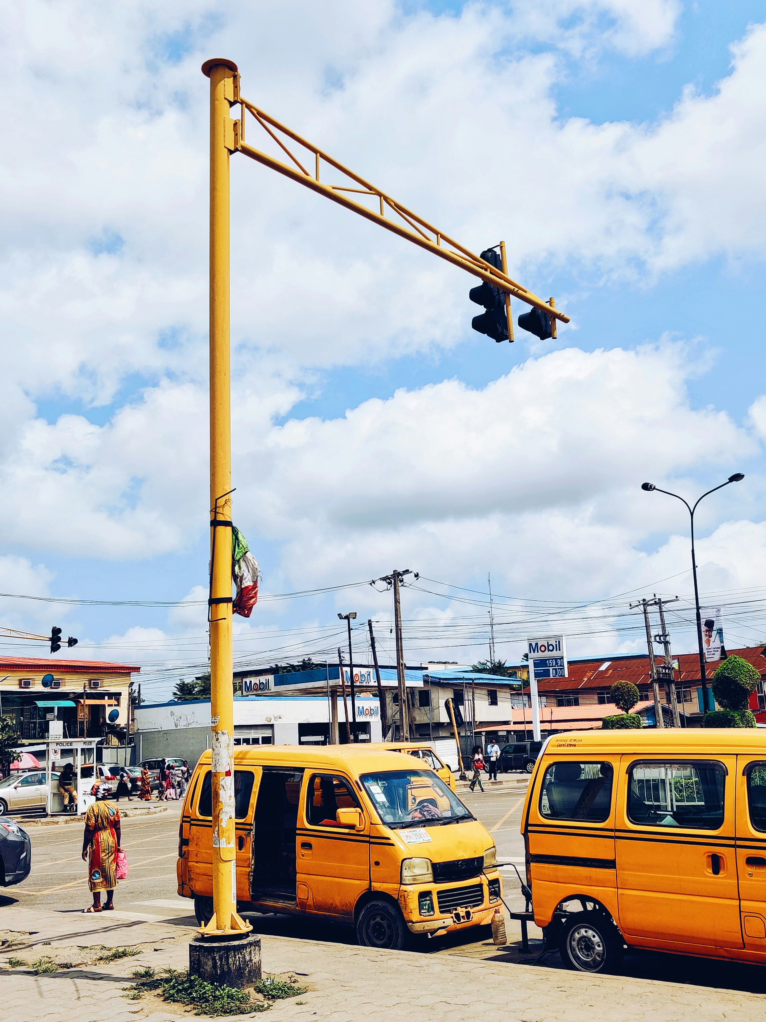 A vibrant urban scene featuring yellow mini-buses and a traffic light against a backdrop of fluffy clouds and commercial buildings.