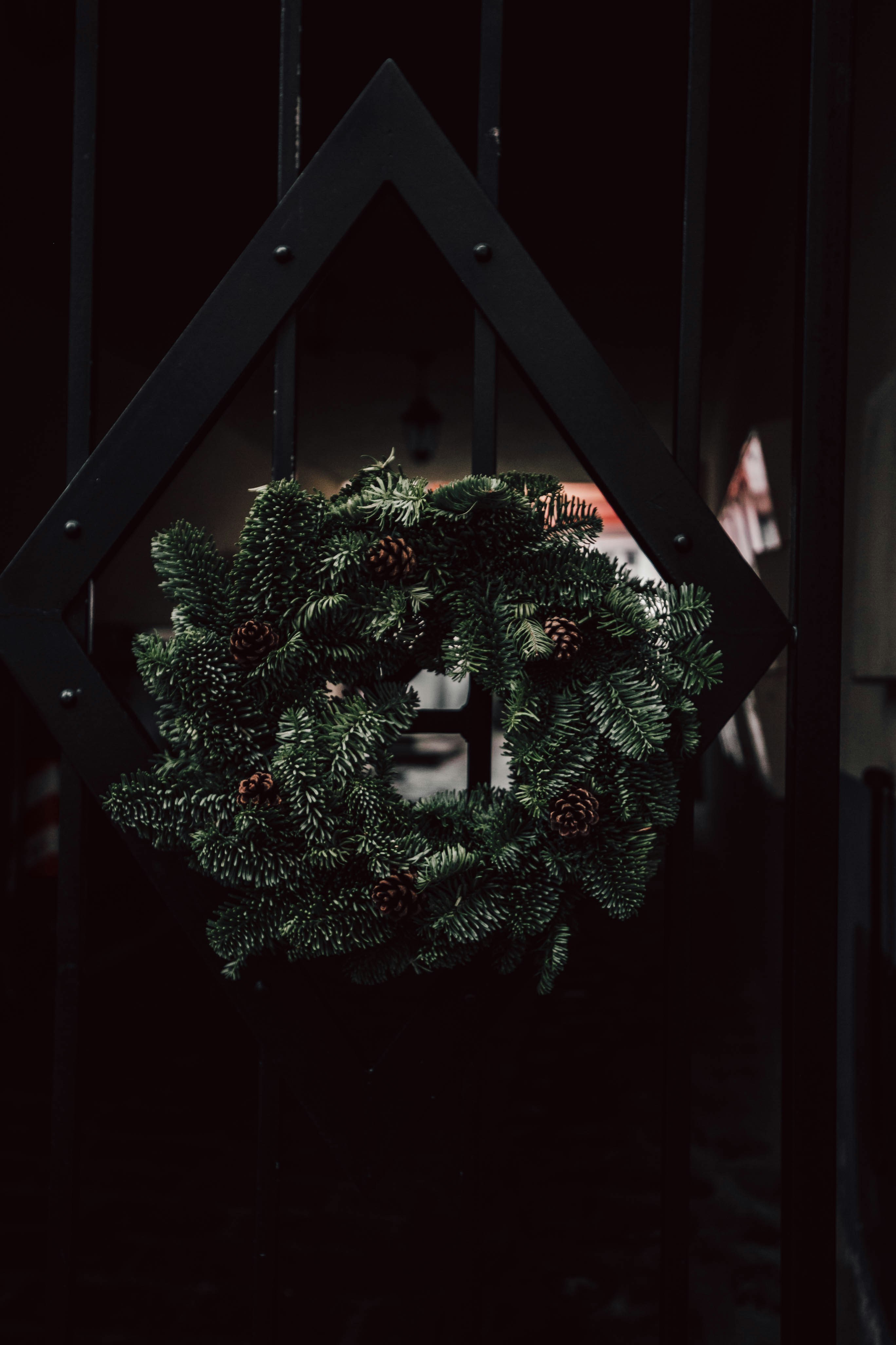 Evergreen wreath adorned with pine cones, hanging on a wrought iron gate. The dark background enhances the wreath's vibrant greens and natural textures.