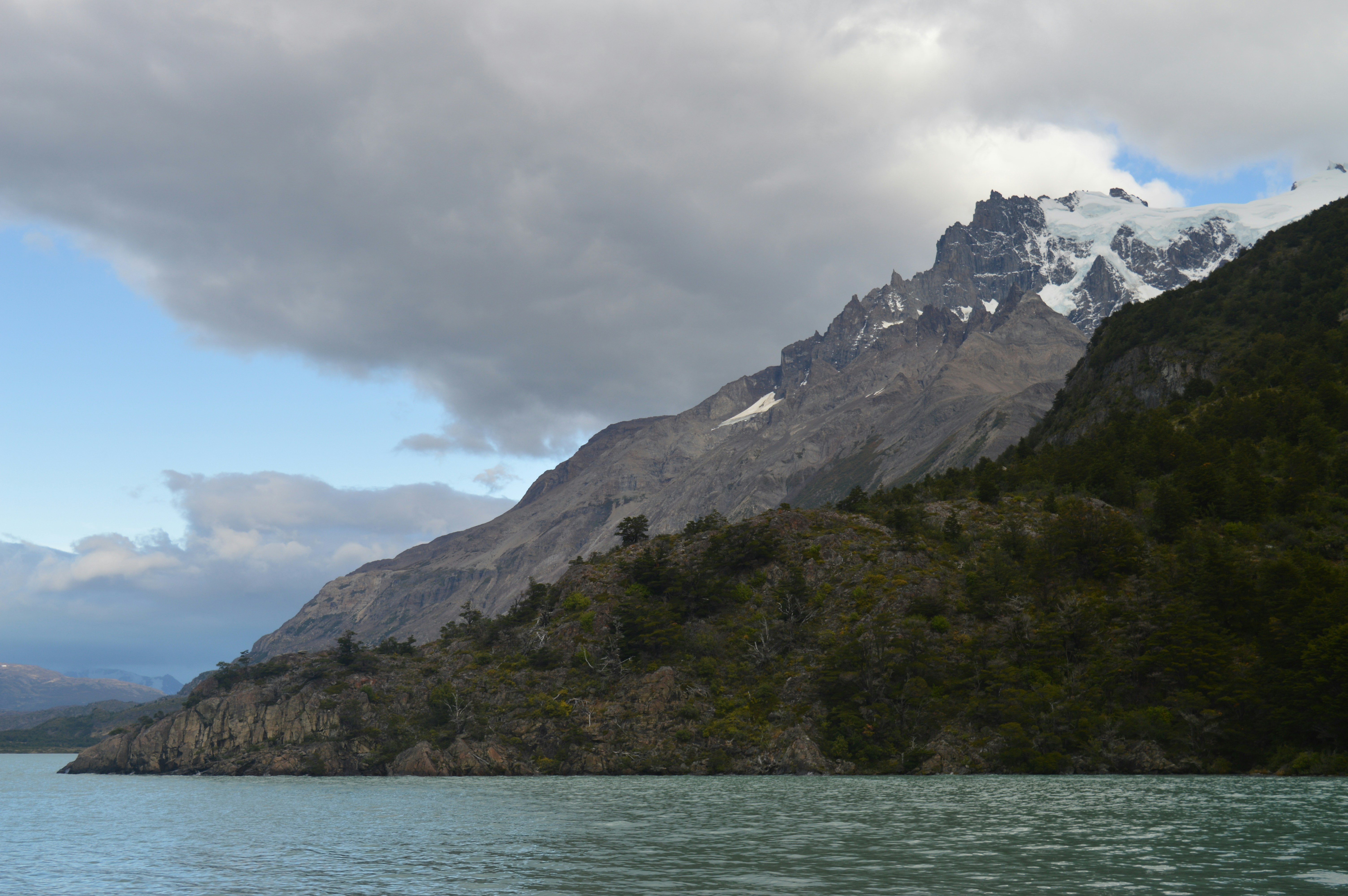 Snow-capped mountains rise dramatically above a tranquil lake, framed by lush greenery along the shoreline.