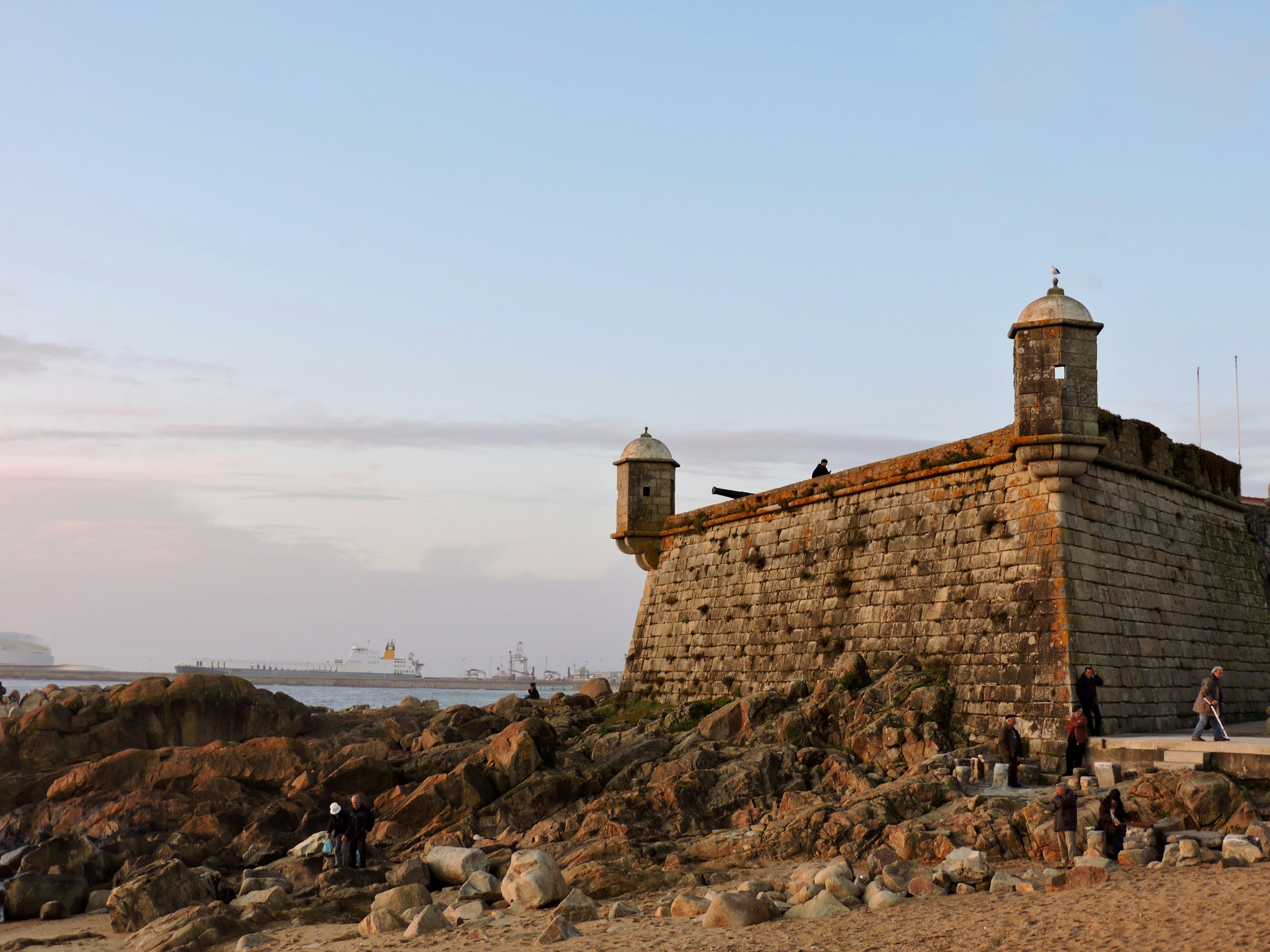 Photograph of a coastal fortress perched on rugged rocks at sunset, with visitors wandering the stone ramparts and the calm sea beyond.