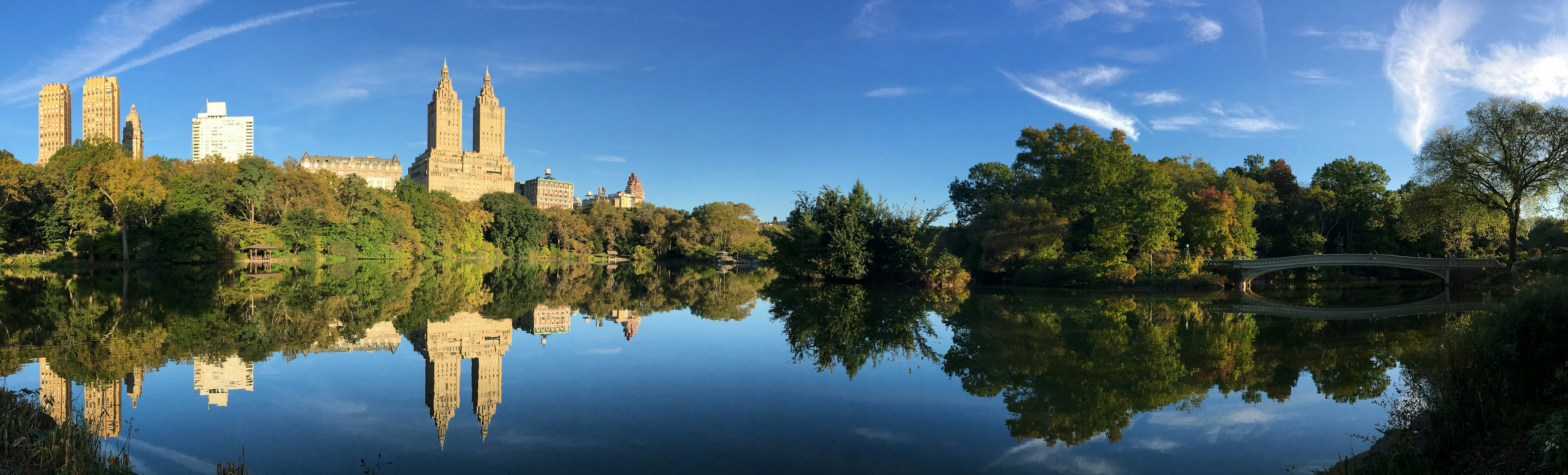 Panoramic view of Central Park showcasing vibrant autumn foliage reflecting in the calm waters, with iconic buildings in the background.