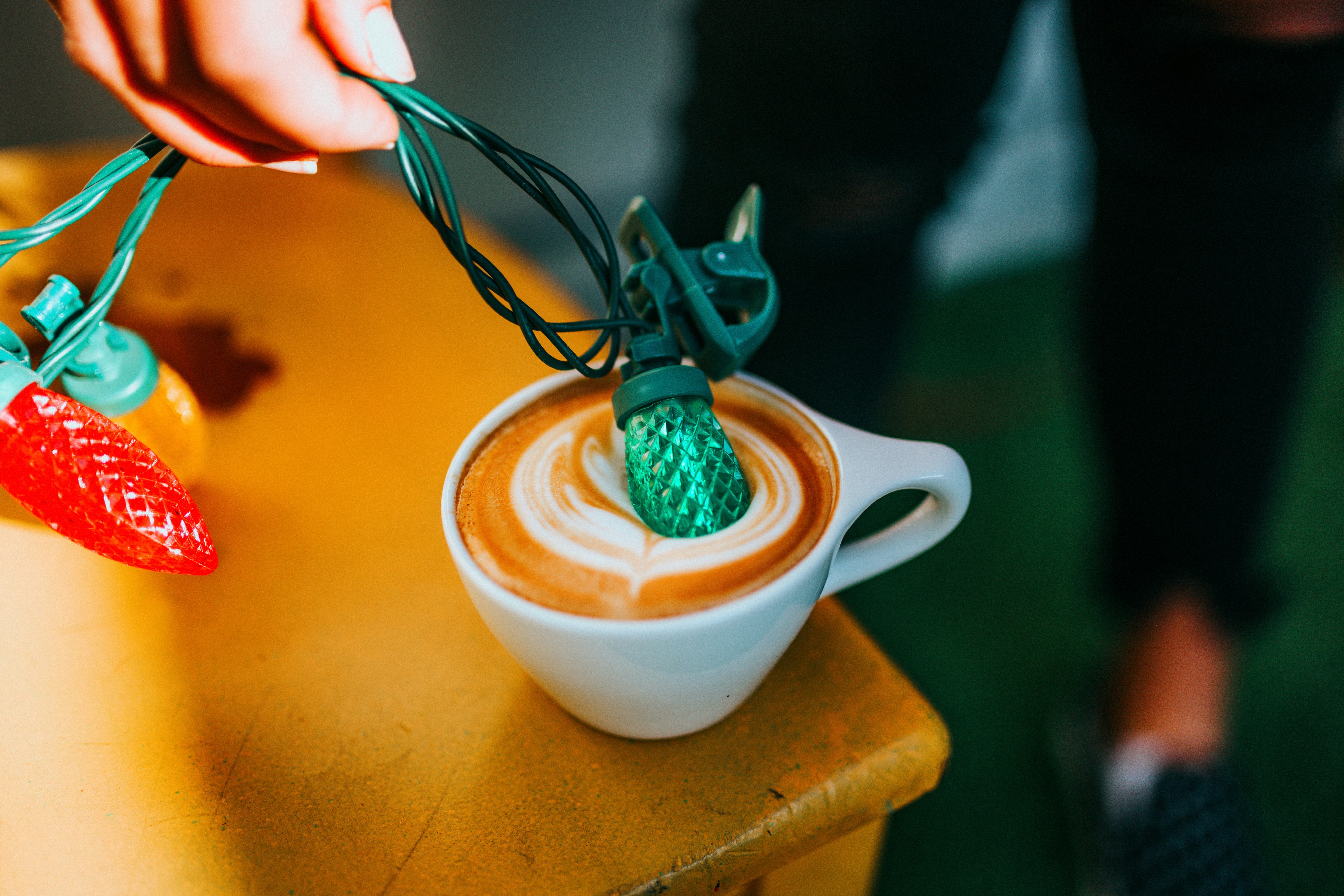 Colorful string lights hover above a latte in a white cup, creating a cheerful ambiance. The scene captures a blend of coffee culture and playful decor.