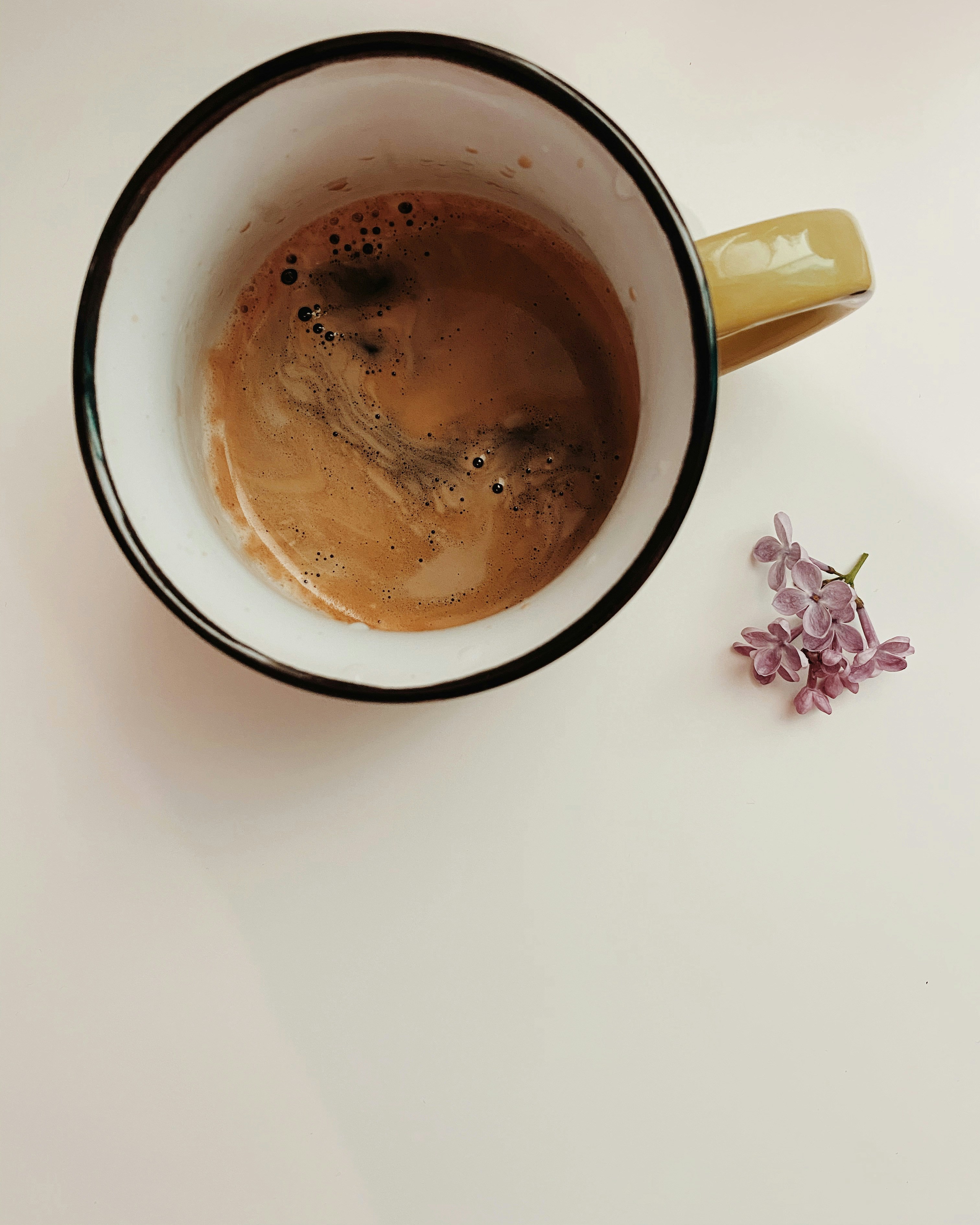 Coffee cup with swirls of crema beside delicate lilac flowers on a minimalist surface.