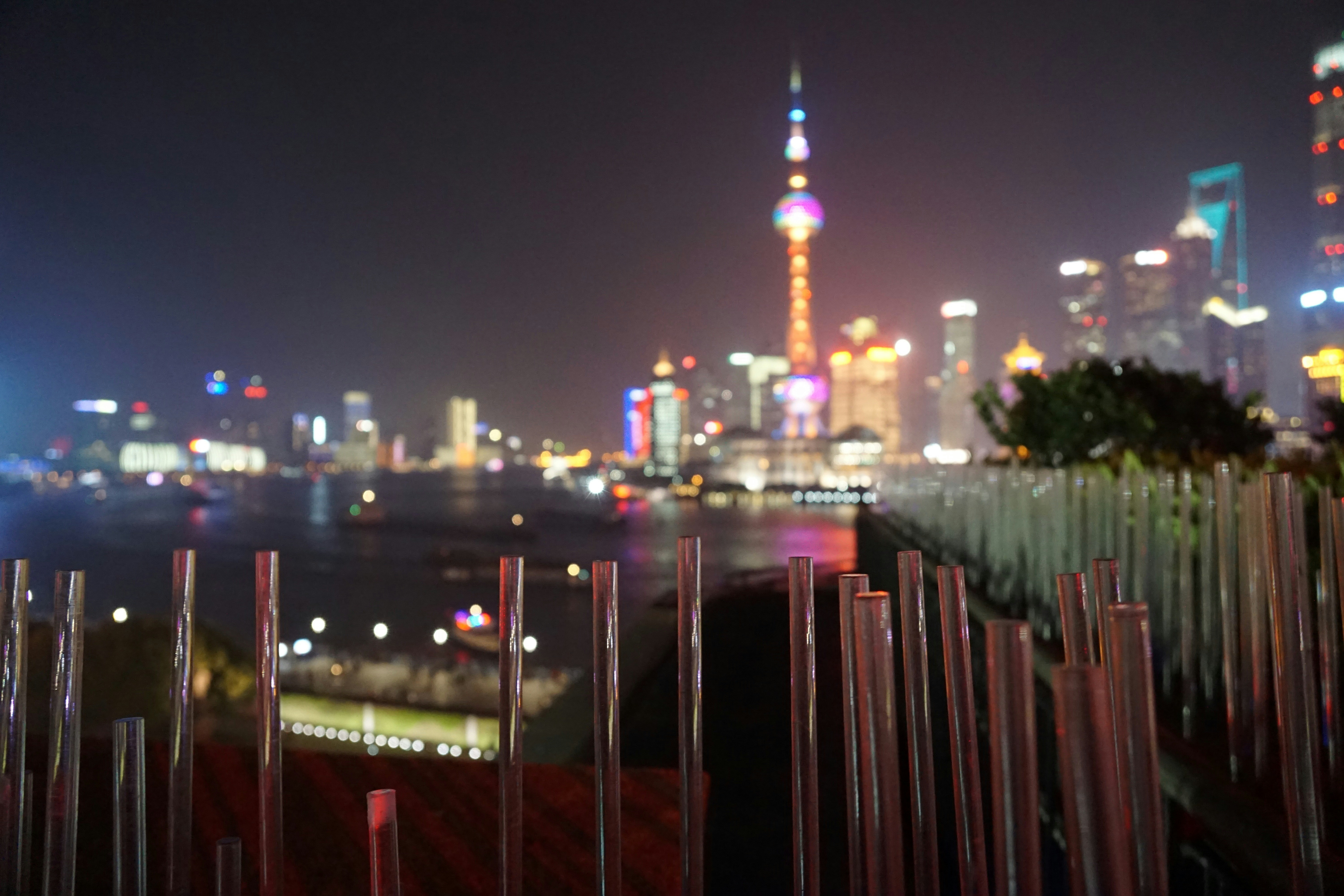 Vertical pieces of wood in the foreground and the lights of Pudong across the river in the background. Soft lavender and pink colors. | city lights during night time