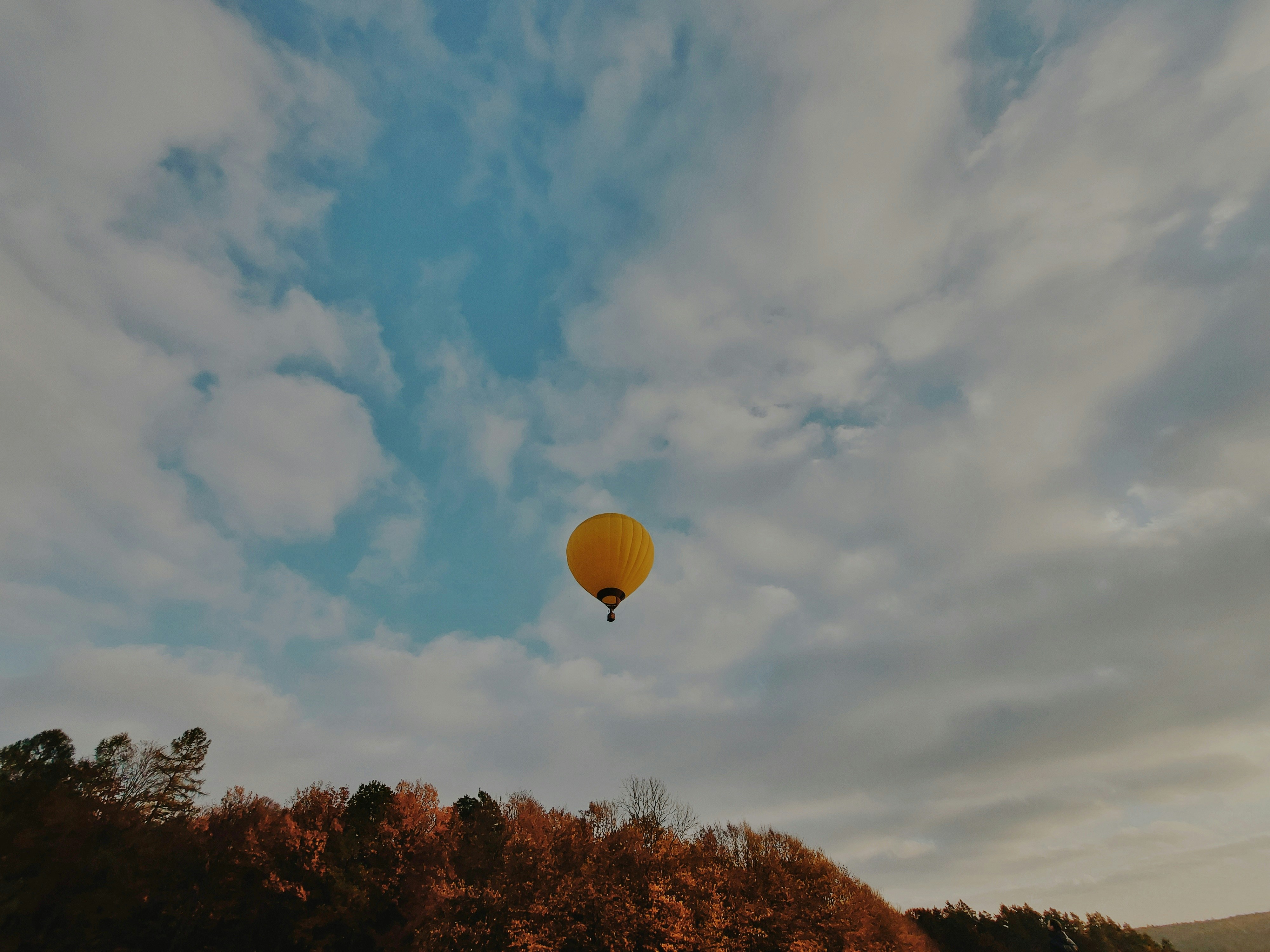 A vibrant yellow hot air balloon floats serenely against a backdrop of wispy clouds and autumn foliage. The scene captures the tranquility of a clear day.