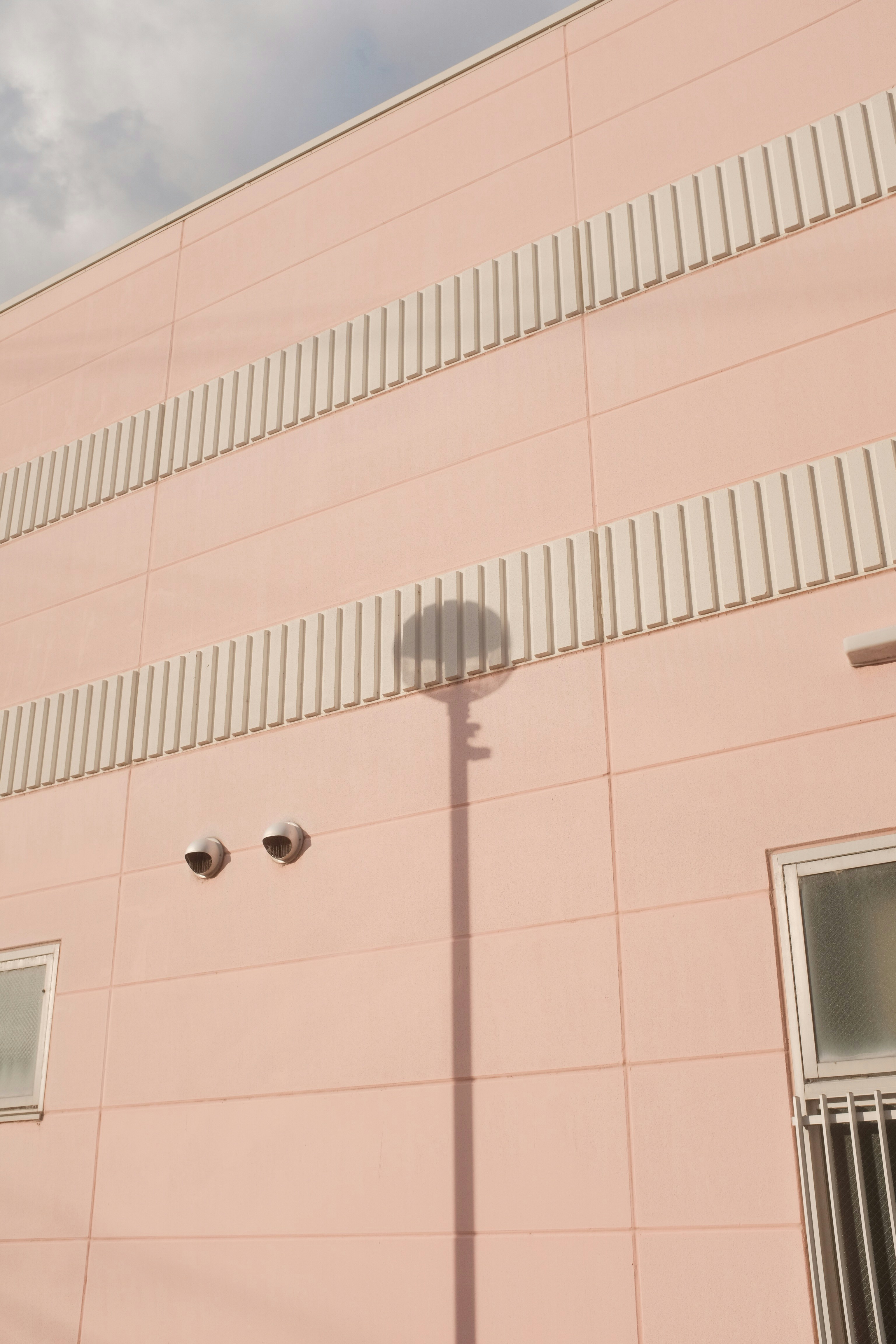 A pale pink building wall with contrasting white horizontal panels, featuring two security cameras and a shadow cast by a streetlight.