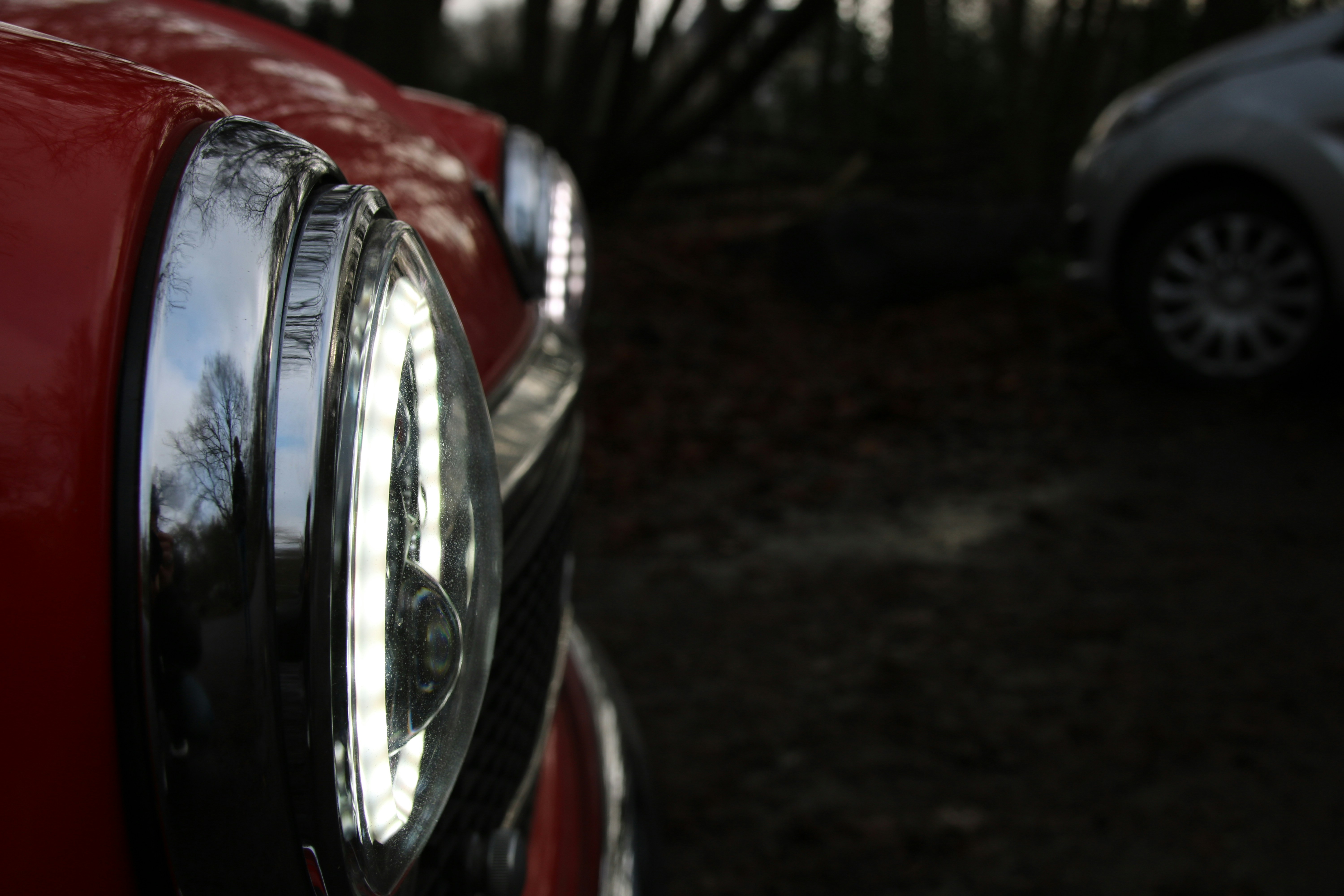 Close-up of a car's headlight, showcasing intricate details and reflections against a blurred background of another vehicle. The warm glow highlights the craftsmanship.