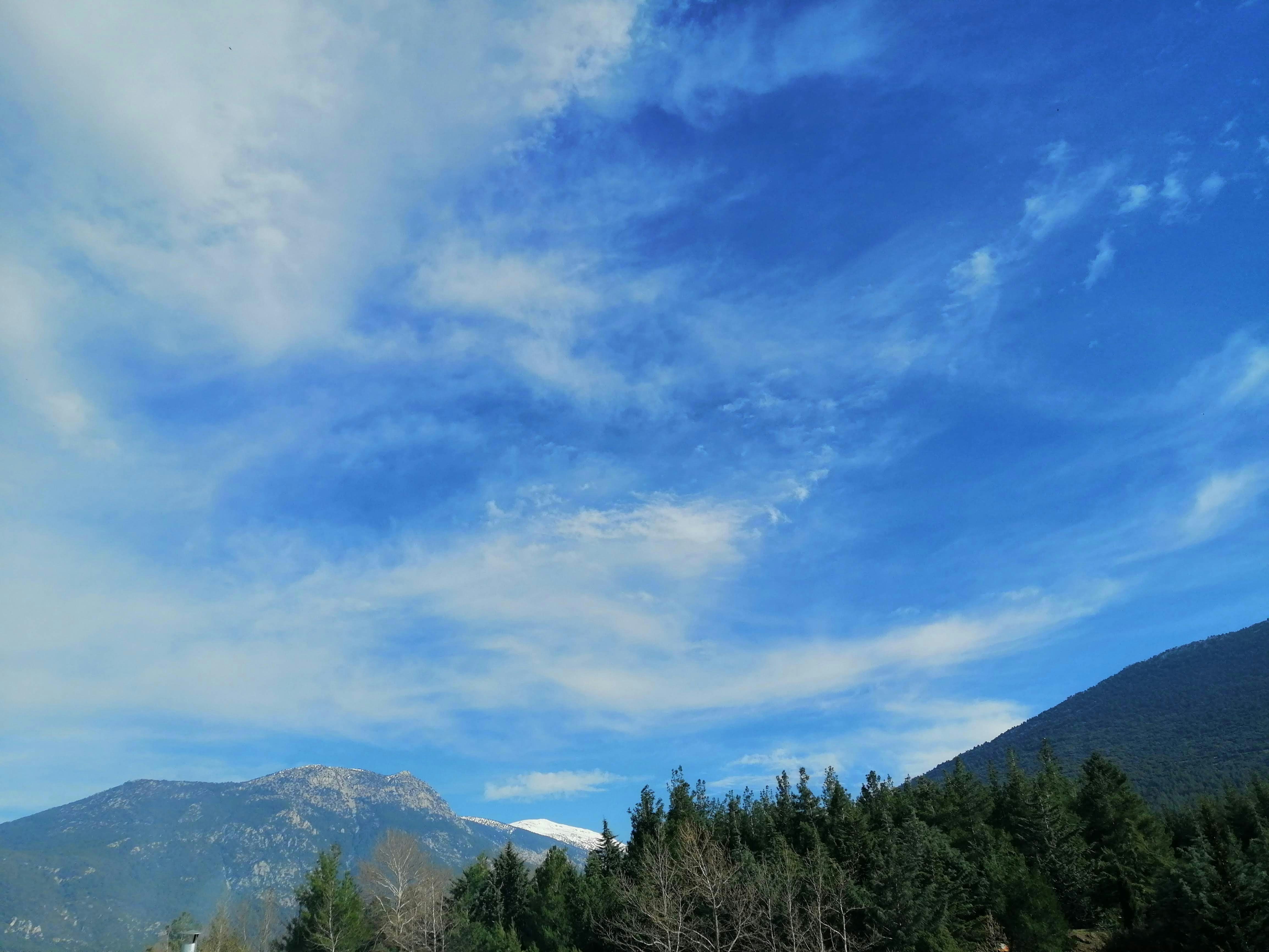 Wide landscape with a vivid blue sky above forested hills. A rocky mountain on the left bears a hint of snow at the peak.
