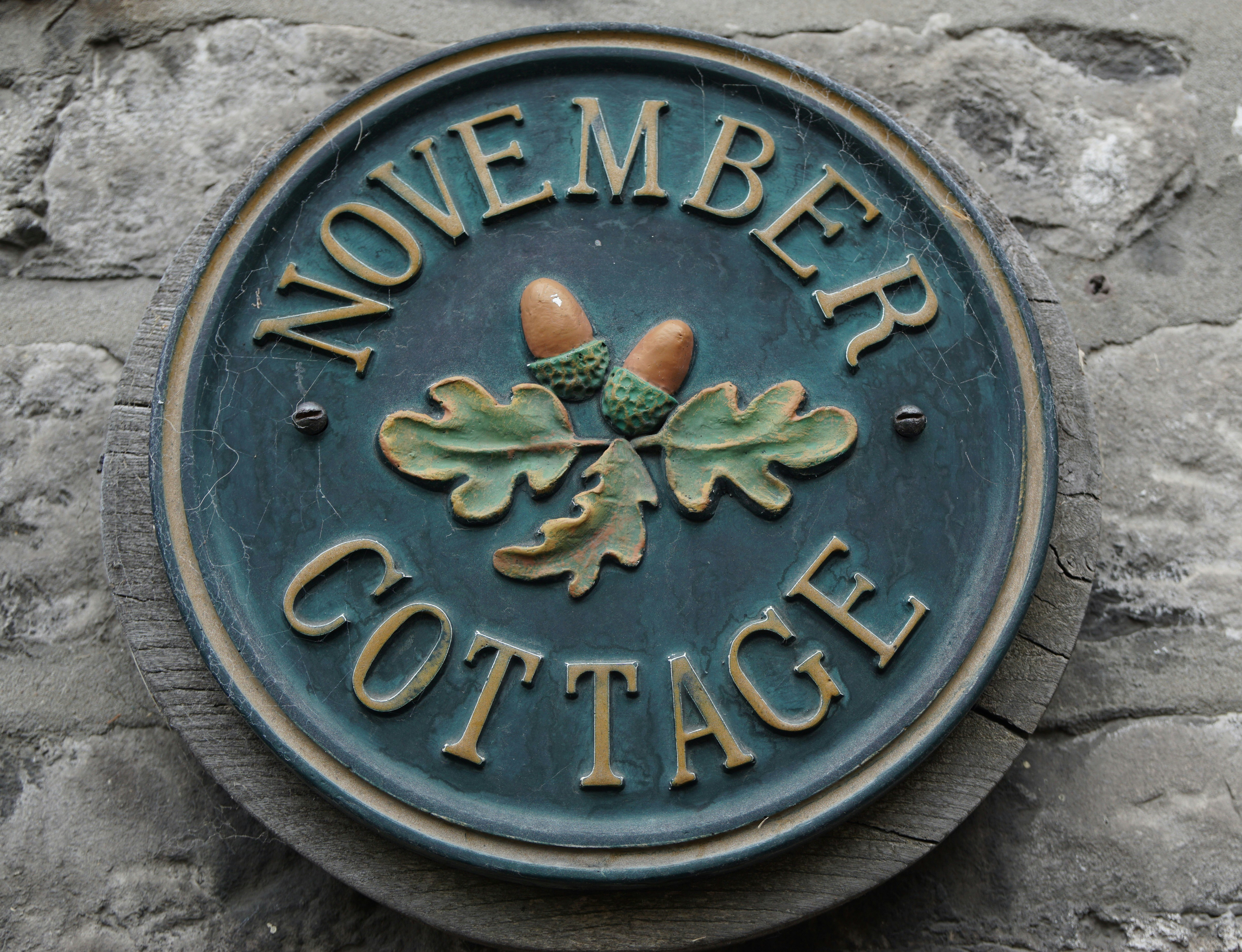 Decorative plaque featuring the words 'November Cottage' with acorns and oak leaves, mounted on a stone wall.