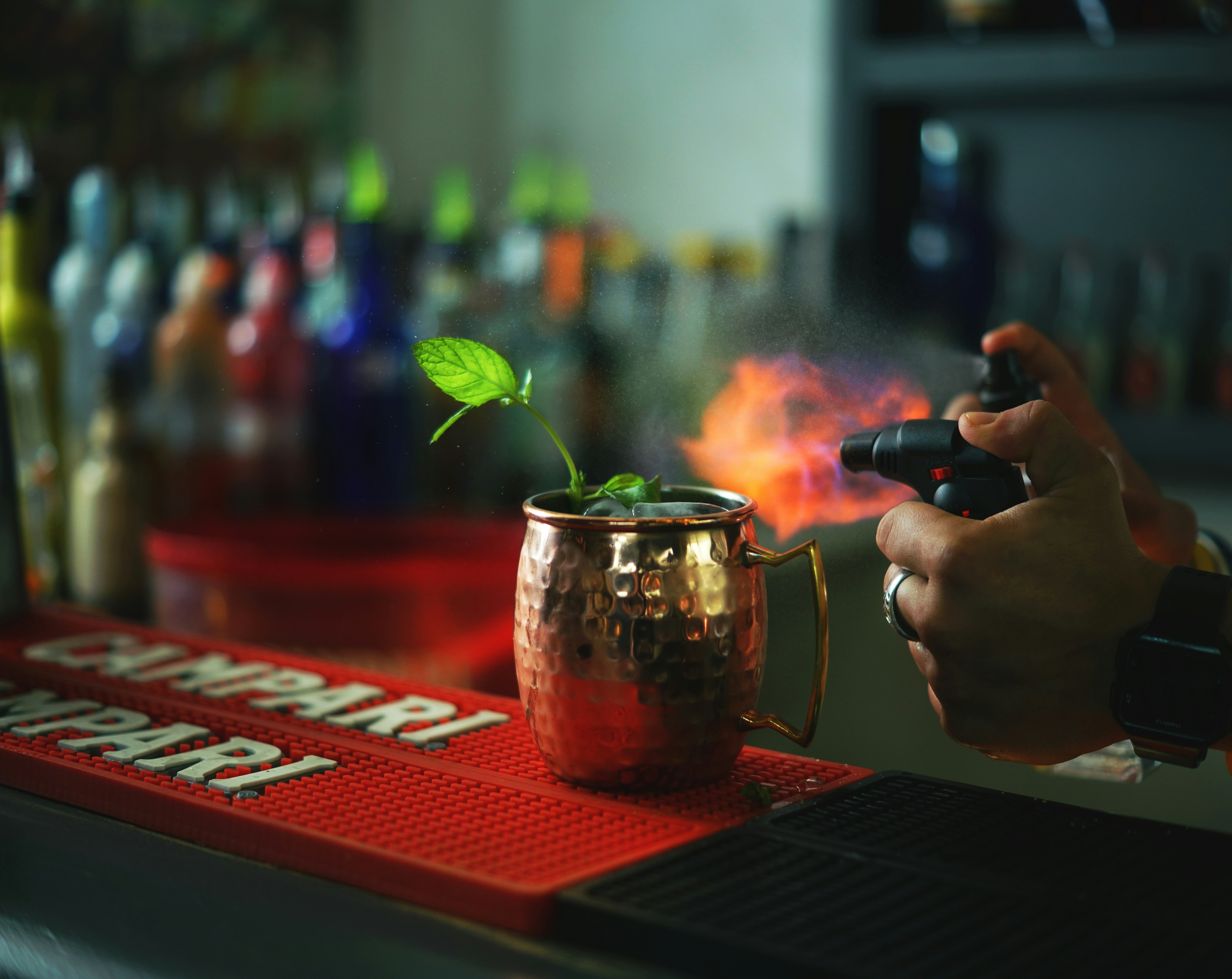 A bartender ignites a sprig of mint above a copper mug, creating an eye-catching display of flames against a blurred bar backdrop.
