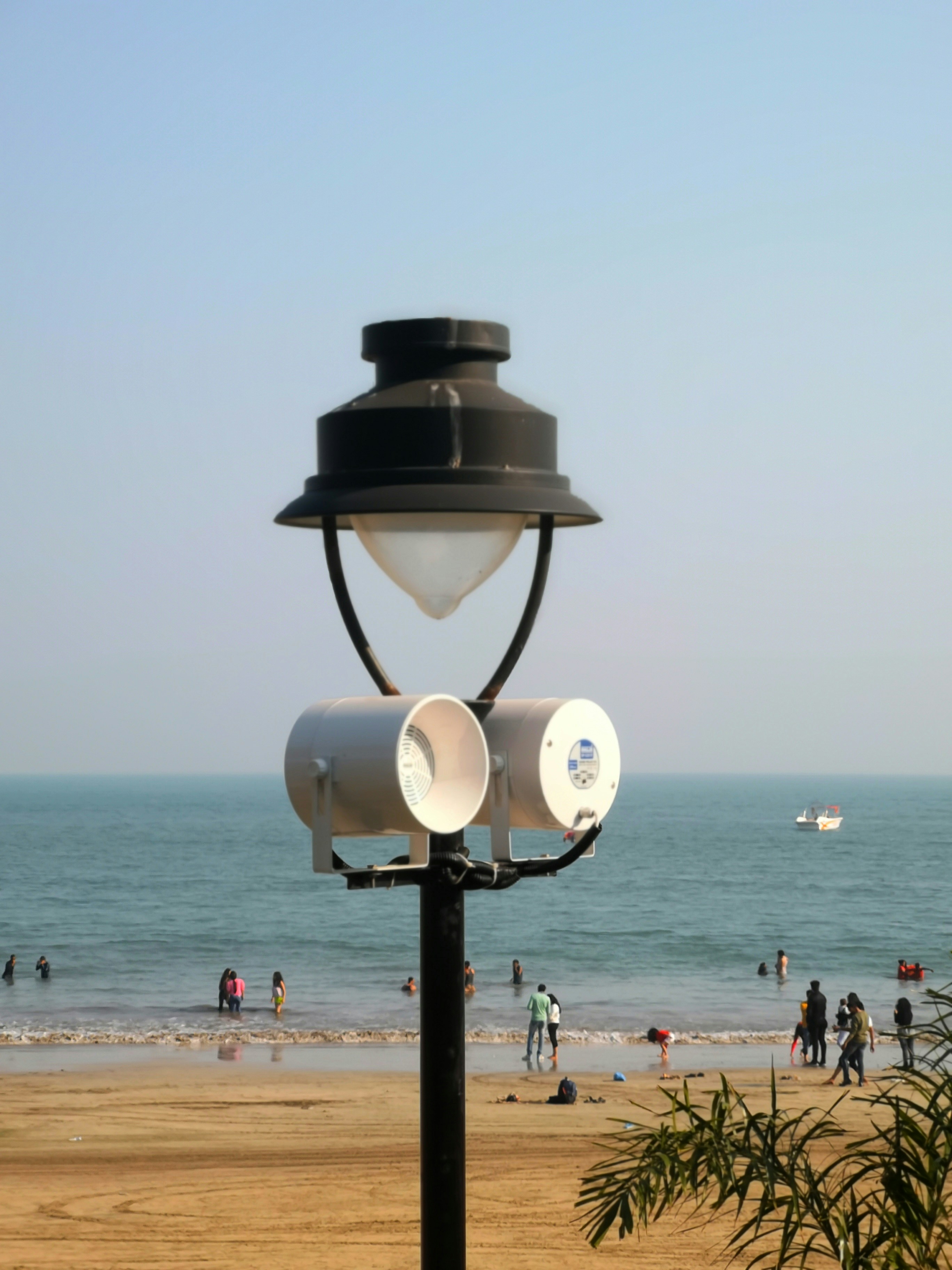 A beachside lamp post with speakers overlooks a sandy shore where people enjoy the water. The gentle waves and distant boat add to the serene atmosphere.