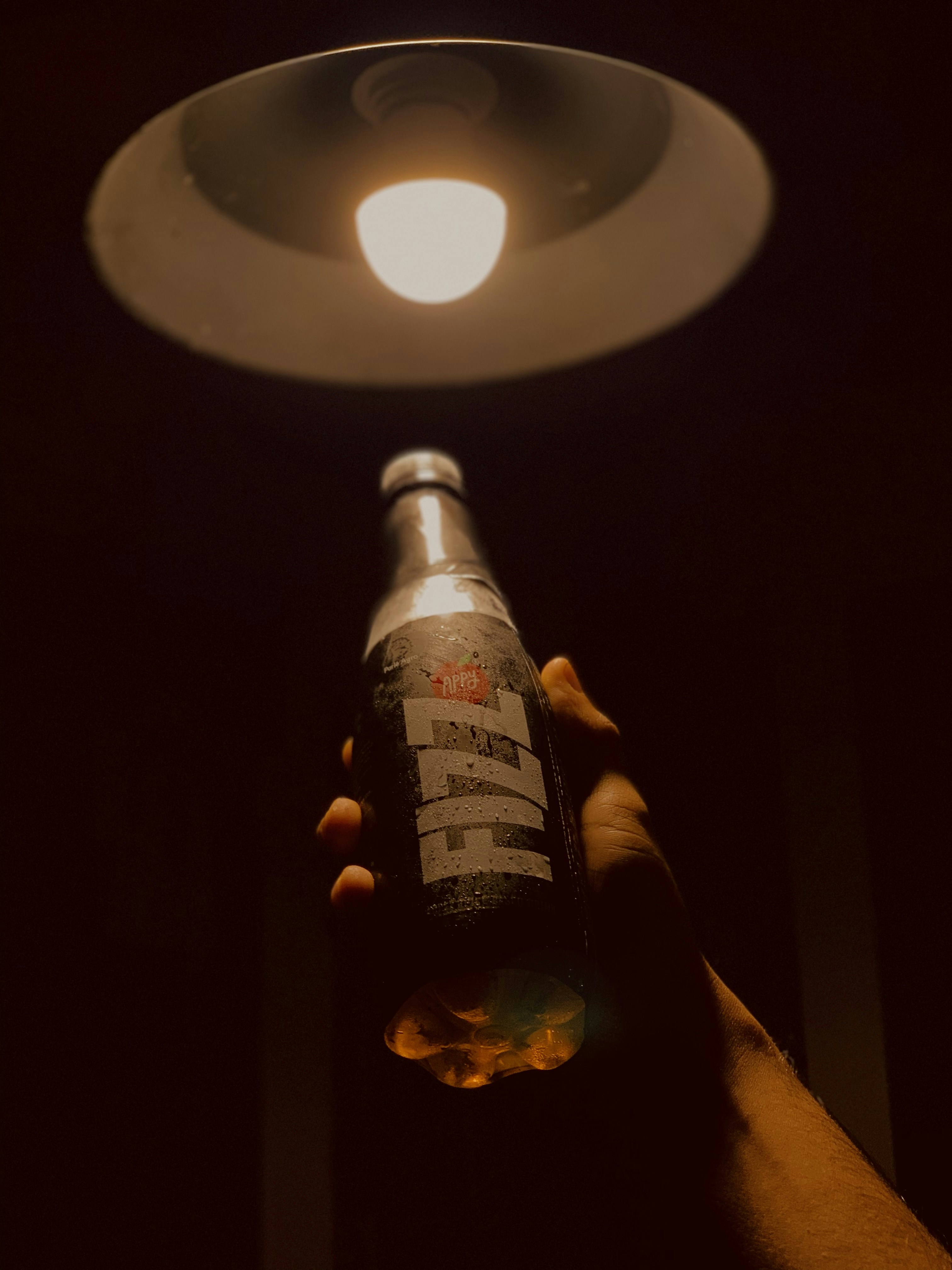 A hand holds a fizzy drink bottle beneath a glowing lamp, creating a warm contrast against a dark background.
