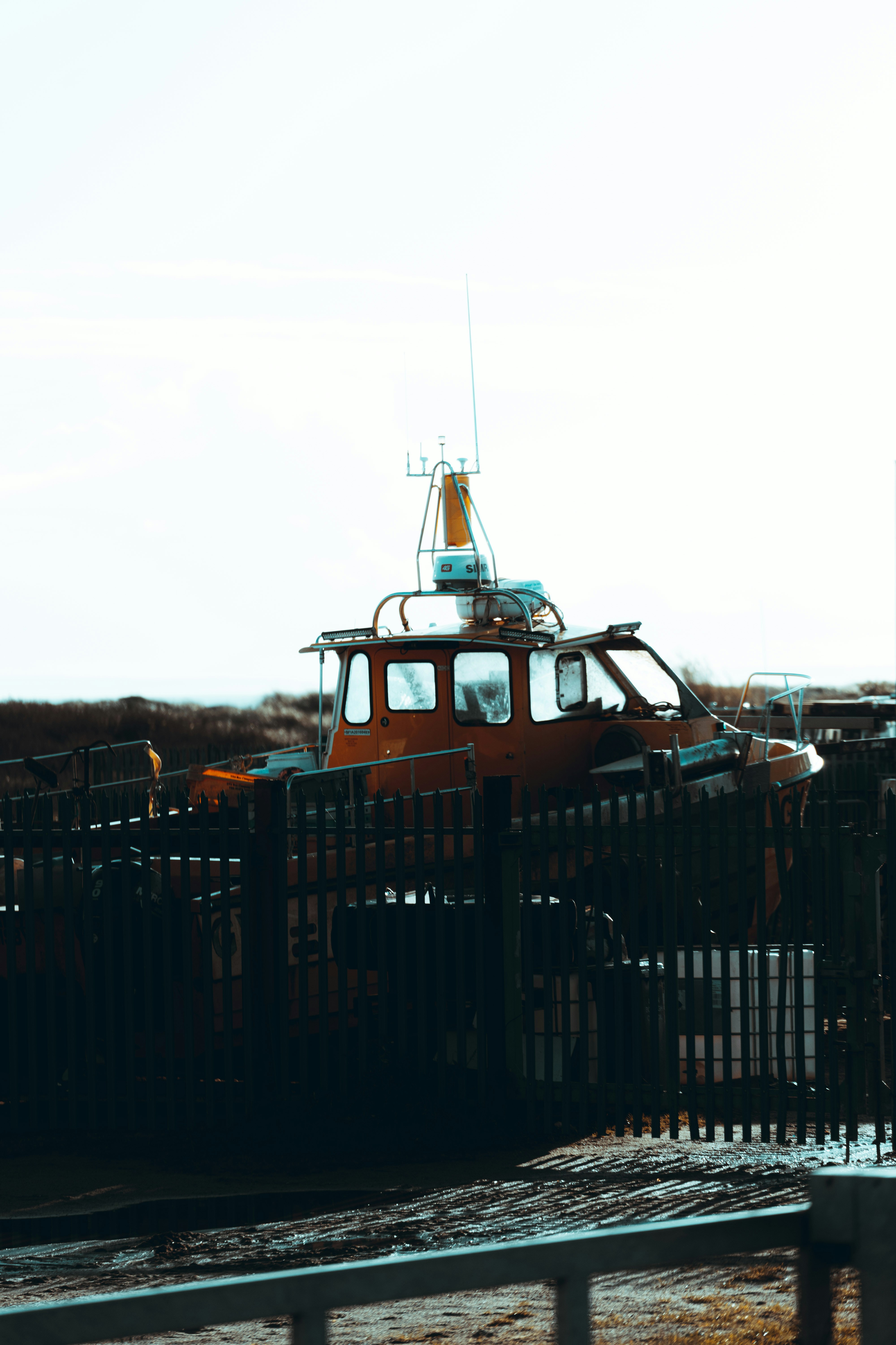 A vibrant orange boat rests against a backdrop of muted colors, framed by a protective fence at a harbor. The scene conveys a sense of calm and readiness.