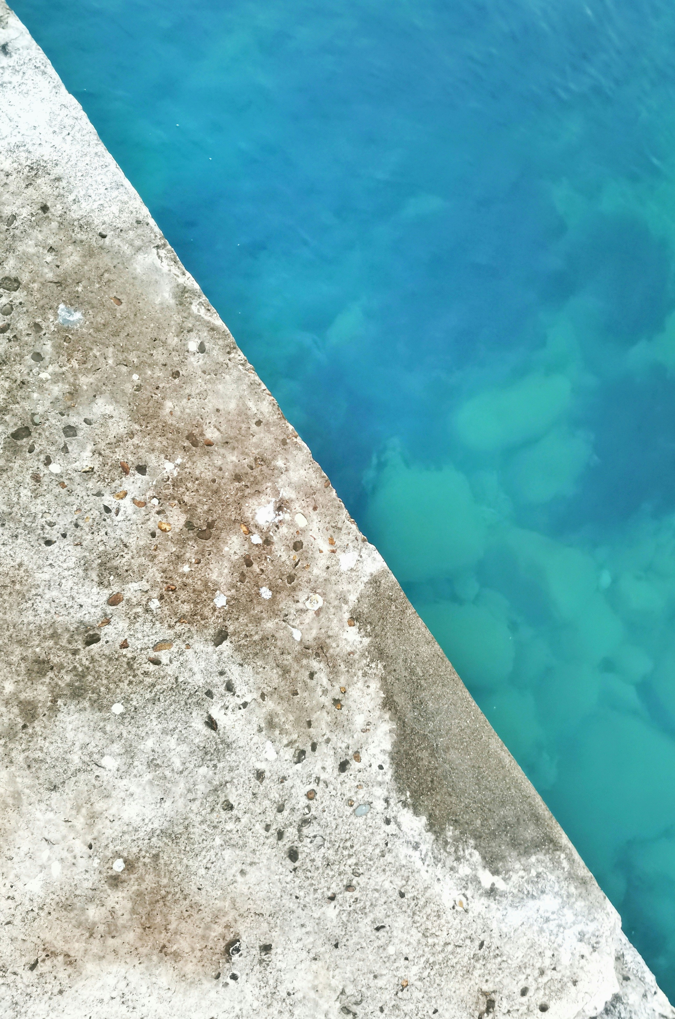 A diagonal concrete edge splits vivid turquoise water, creating bold color and texture contrast. The composition emphasizes the boundary between solid material and liquid depth.