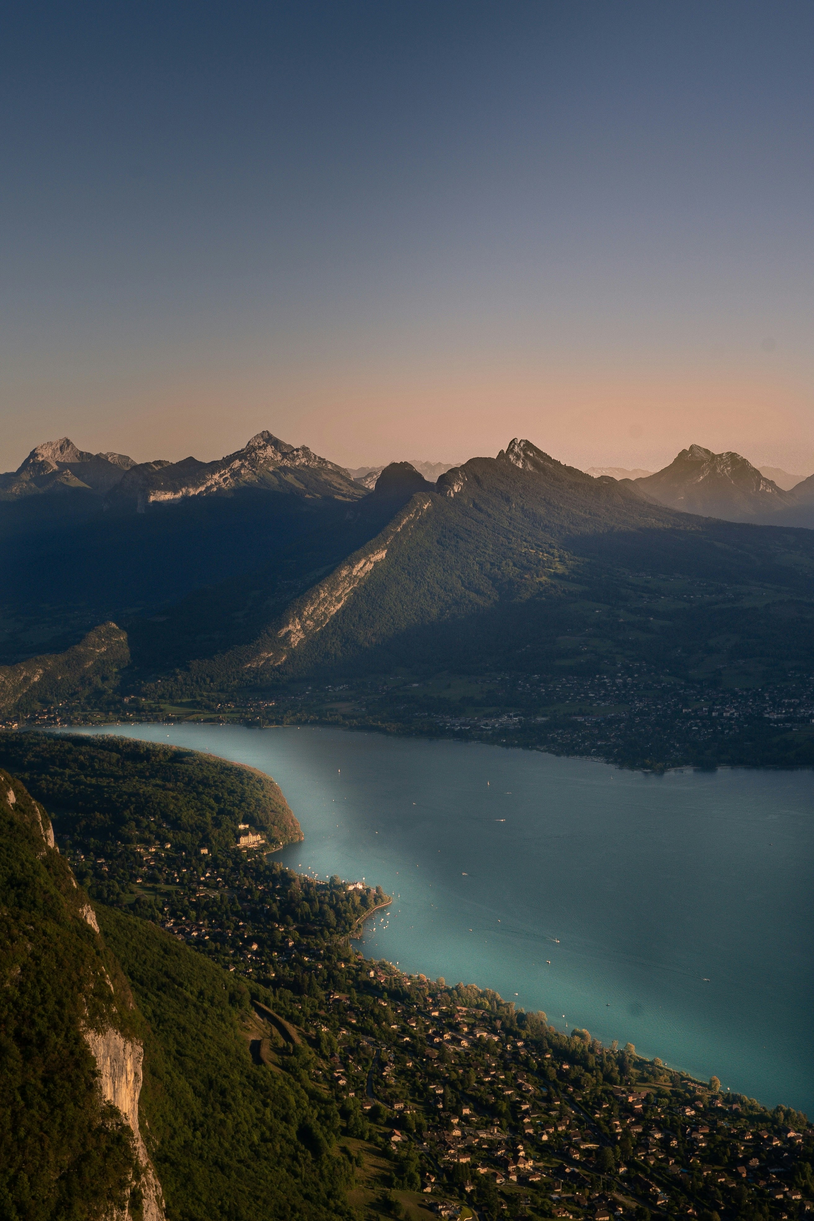 Aerial view of a tranquil lake surrounded by lush greenery and majestic mountains at dusk.
