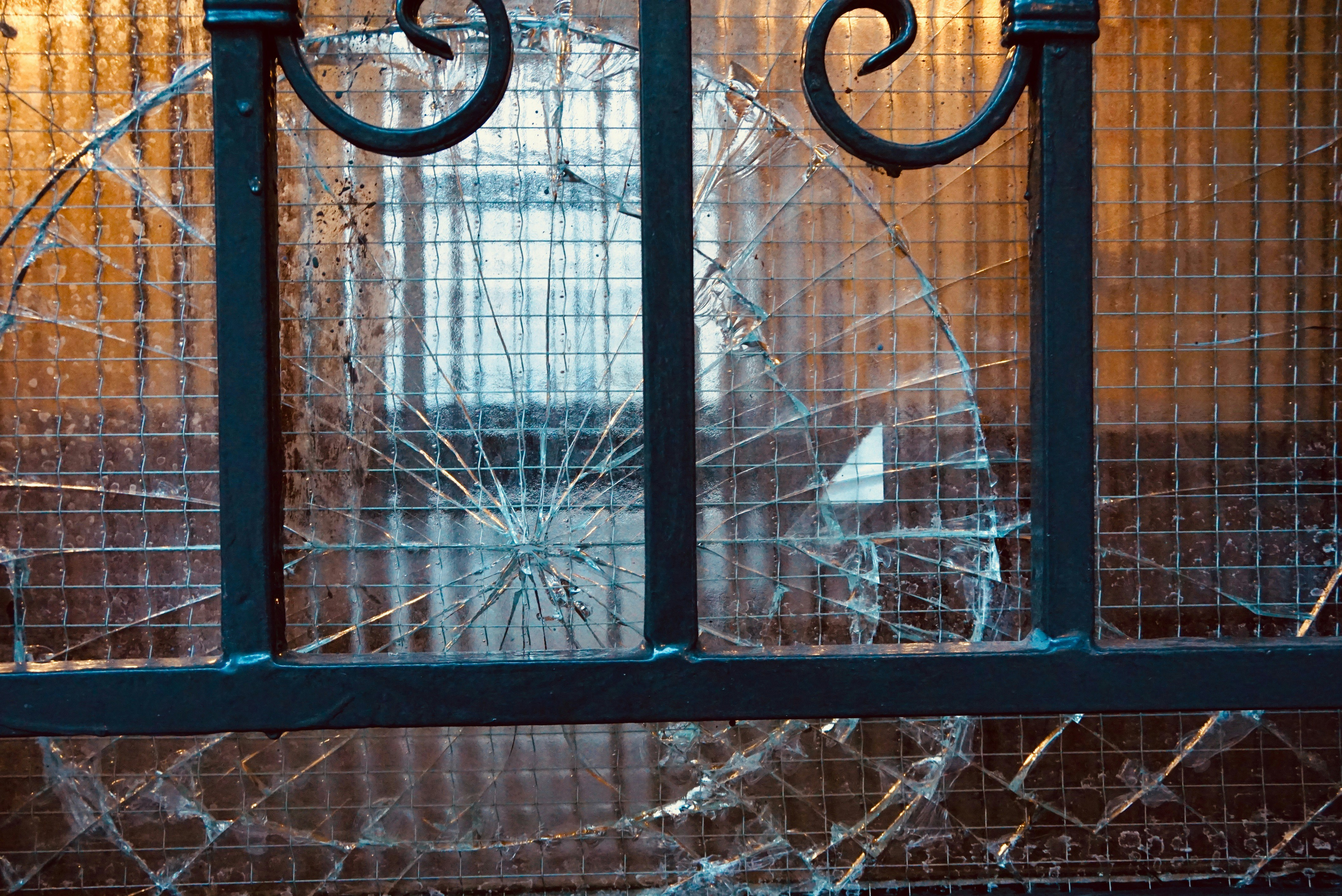Broken glass door with intricate metalwork, revealing a blurred interior. The shattered pattern creates a striking contrast against the warm background.