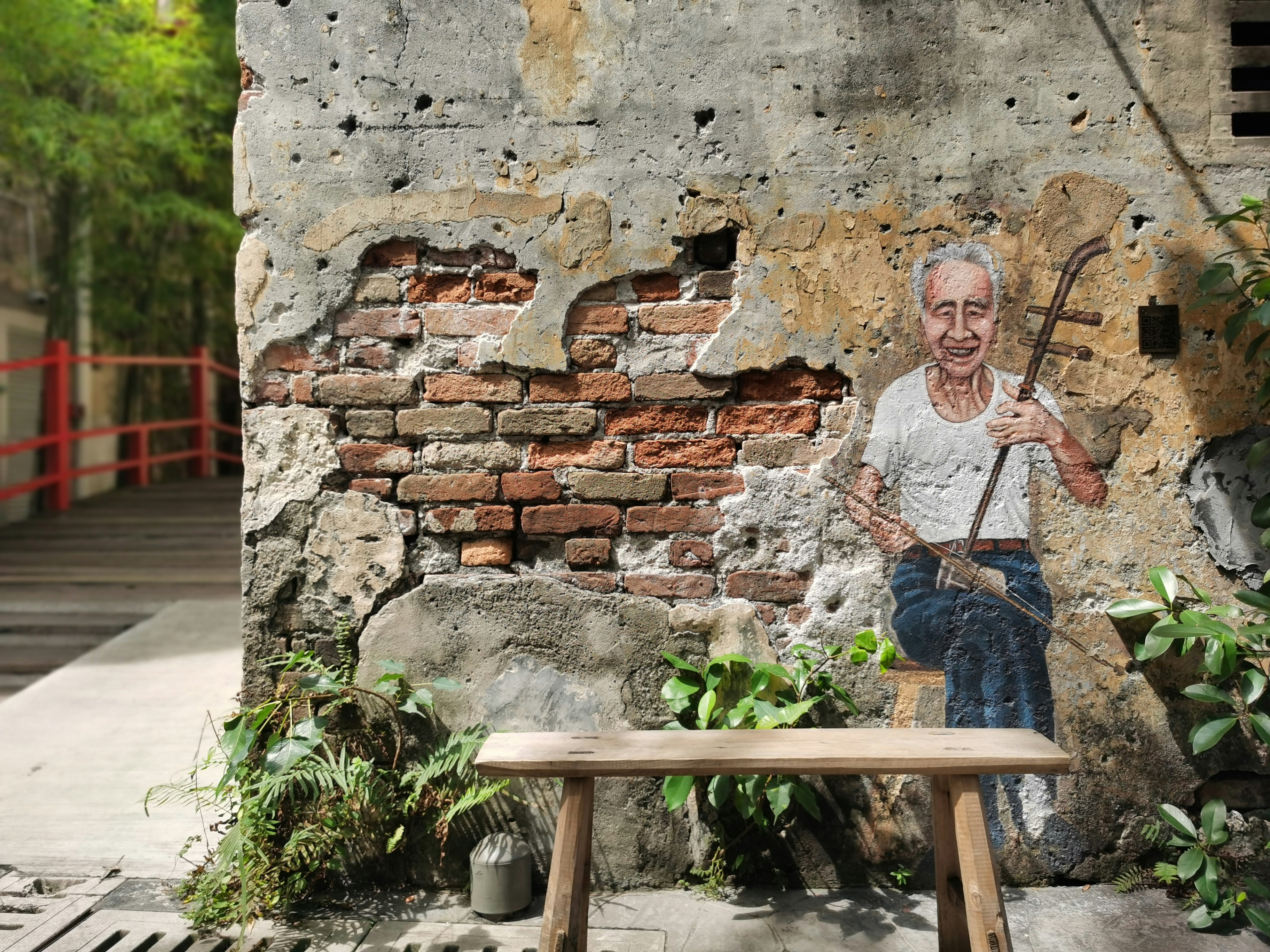Mural painting of an elderly man playing a bowed string instrument on a weathered brick wall beside a wooden bench and greenery.