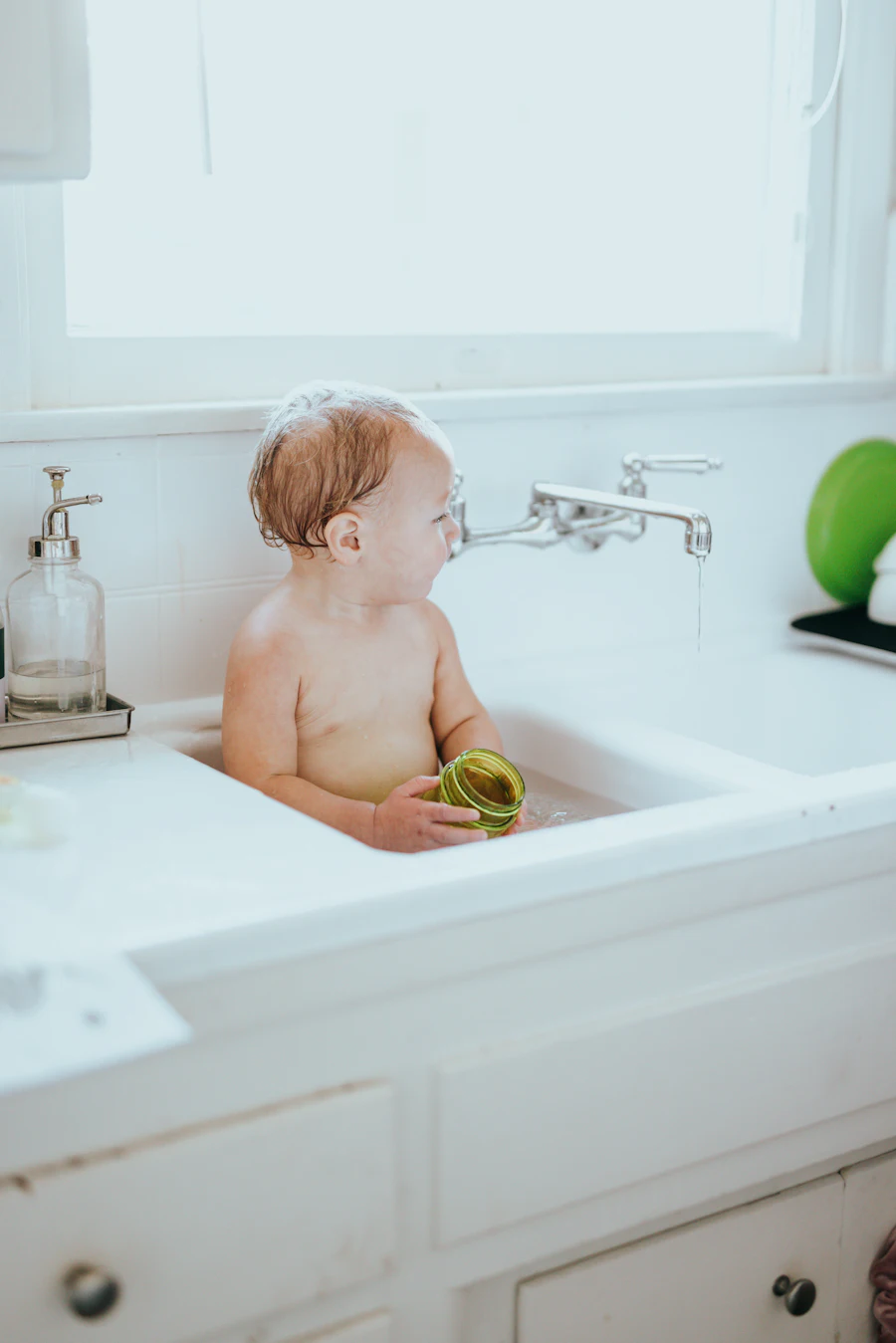 Young toddler sits in a white kitchen sink, washing a green cup under a bright window.