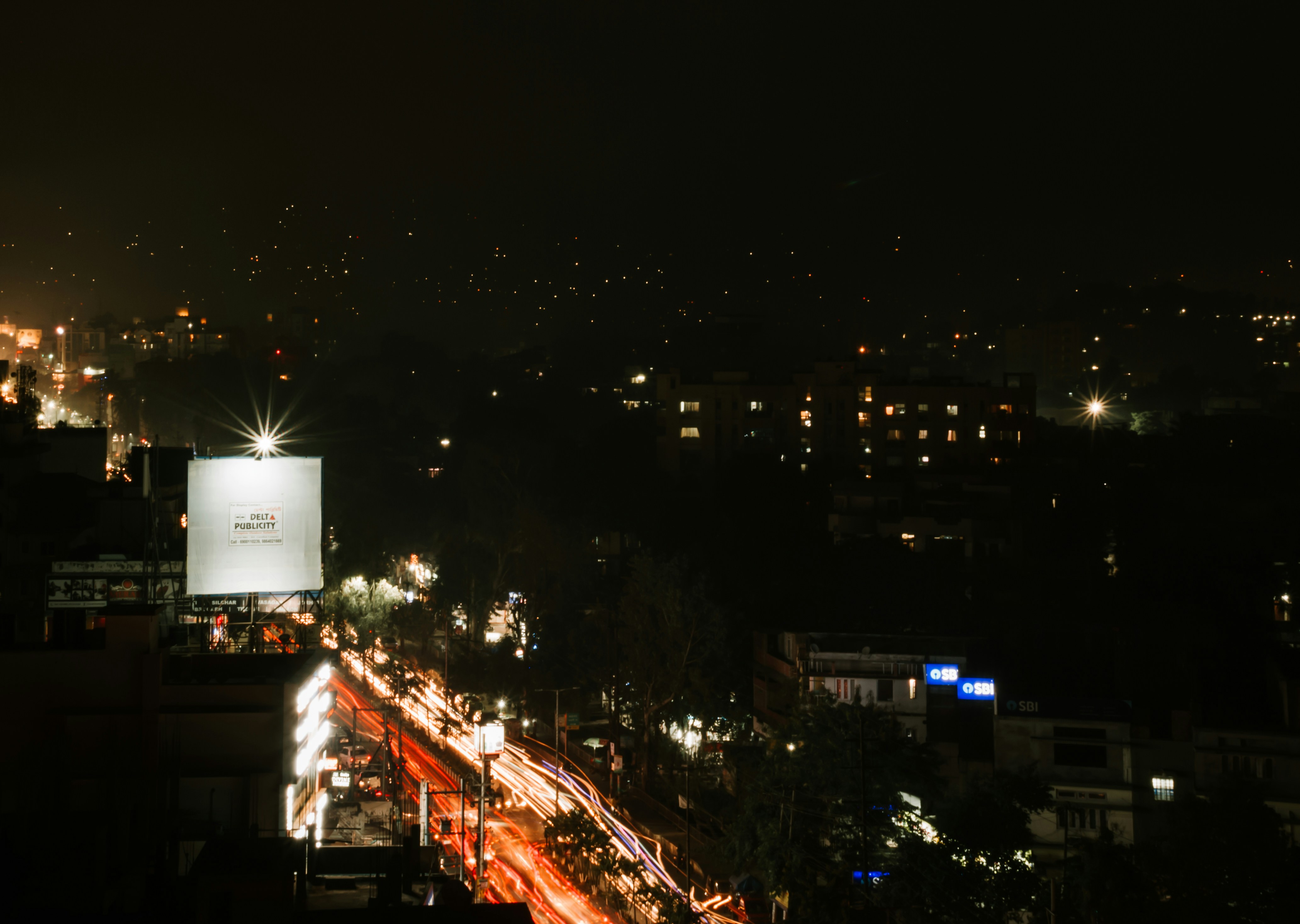 Cityscape at night with streaks of car lights on a busy road and scattered building lights under a dark sky.