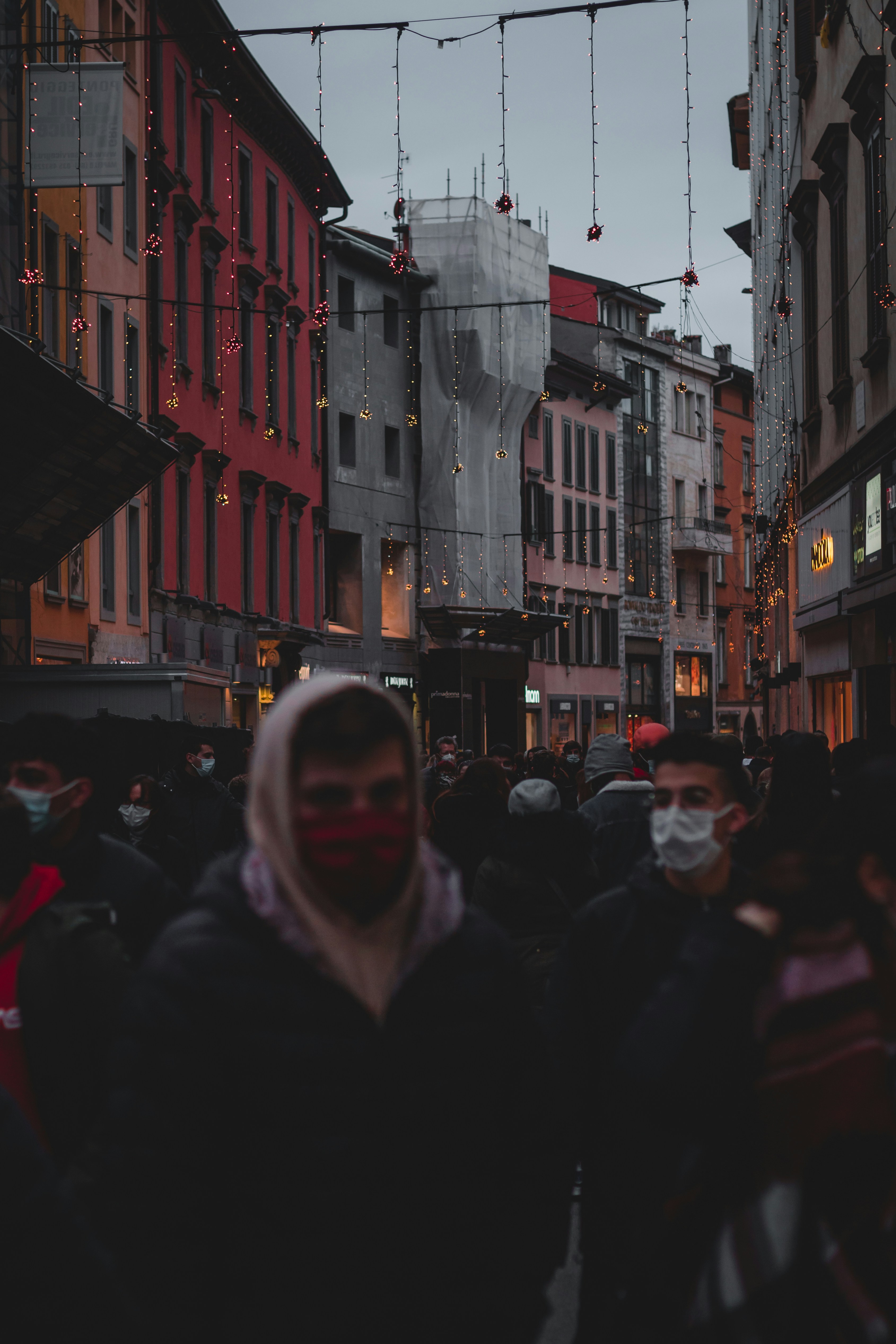 Crowd of people wearing masks walking through a narrow street adorned with hanging lights during dusk.