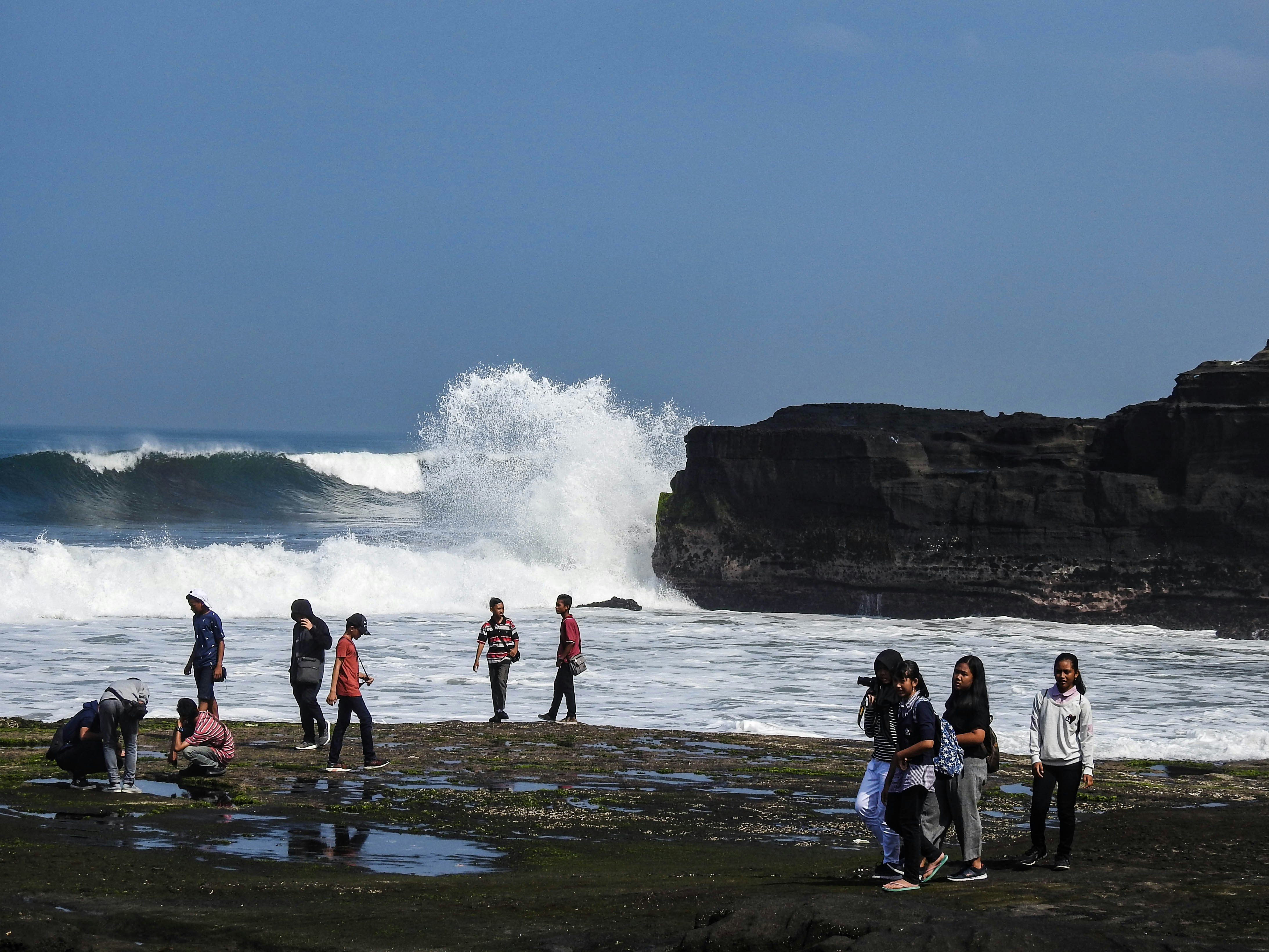 Groups of people exploring rocky tidal pools as waves crash against the shore under a clear blue sky.