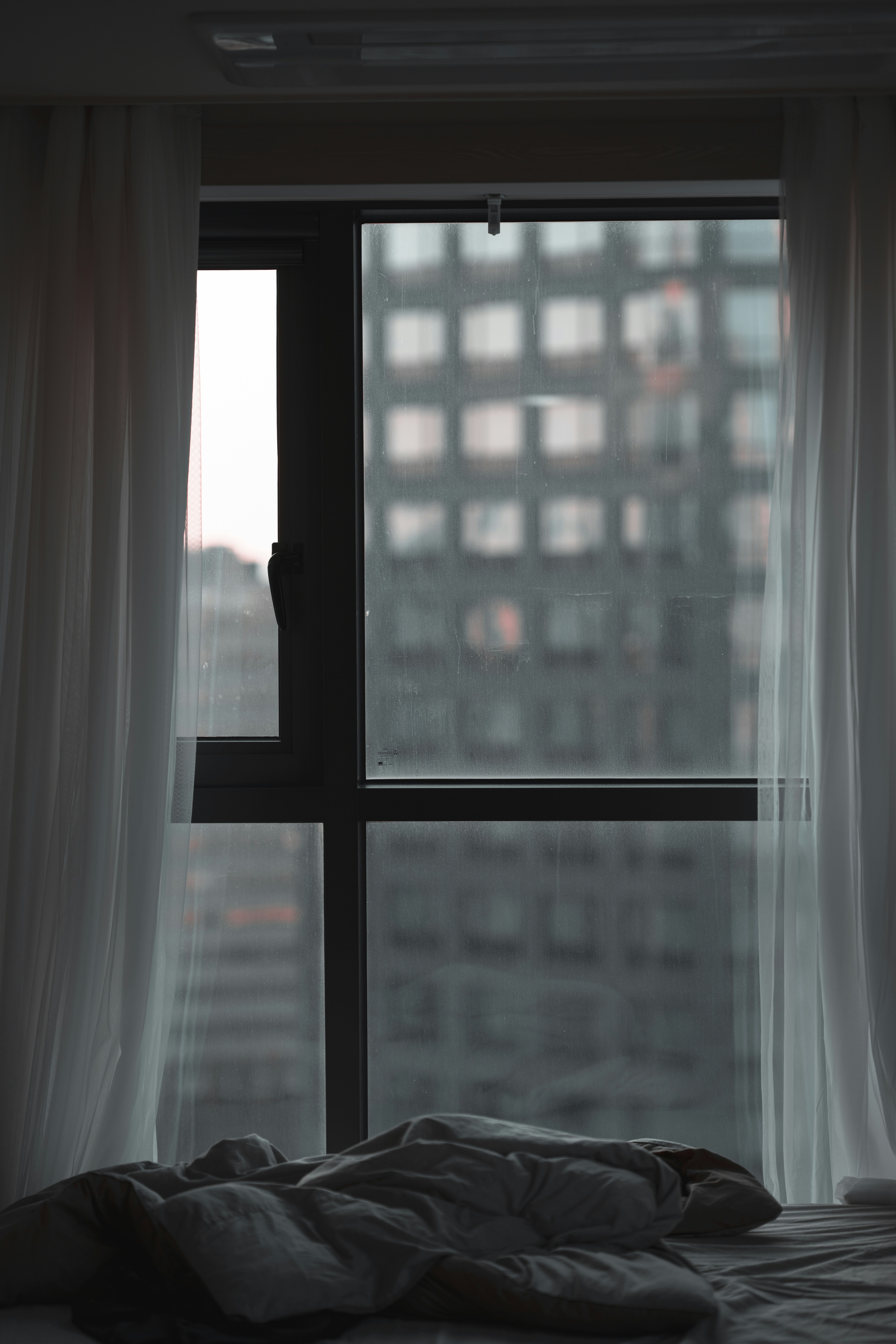 Translucent curtains frame a window overlooking a cityscape, blending indoor comfort with urban life. The soft folds of bedding suggest a moment of pause amidst the bustling backdrop.