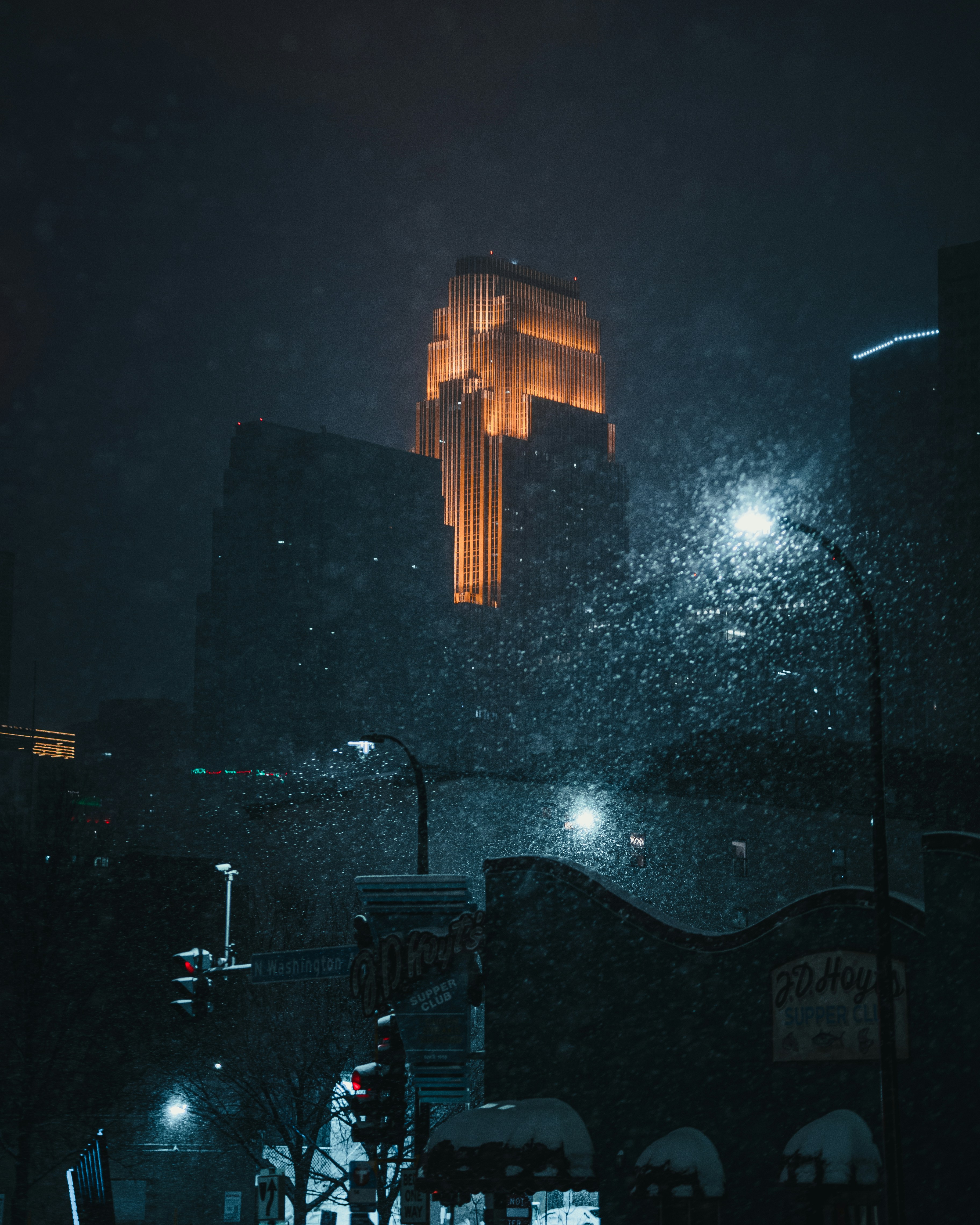 A towering skyscraper glows warmly against a backdrop of swirling snowflakes in a nighttime urban setting.