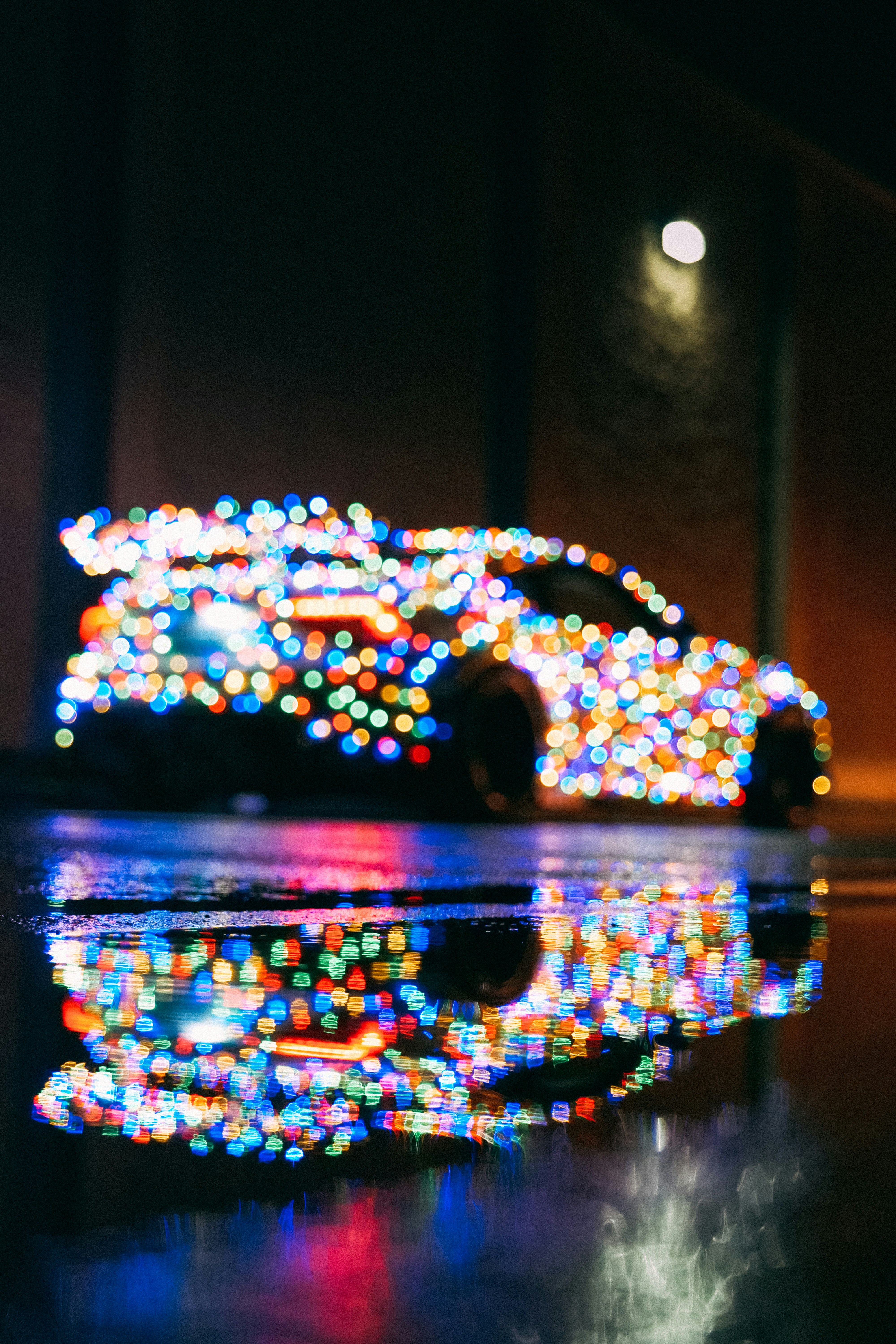 A car adorned with colorful holiday lights, reflecting in a wet surface under dim lighting.