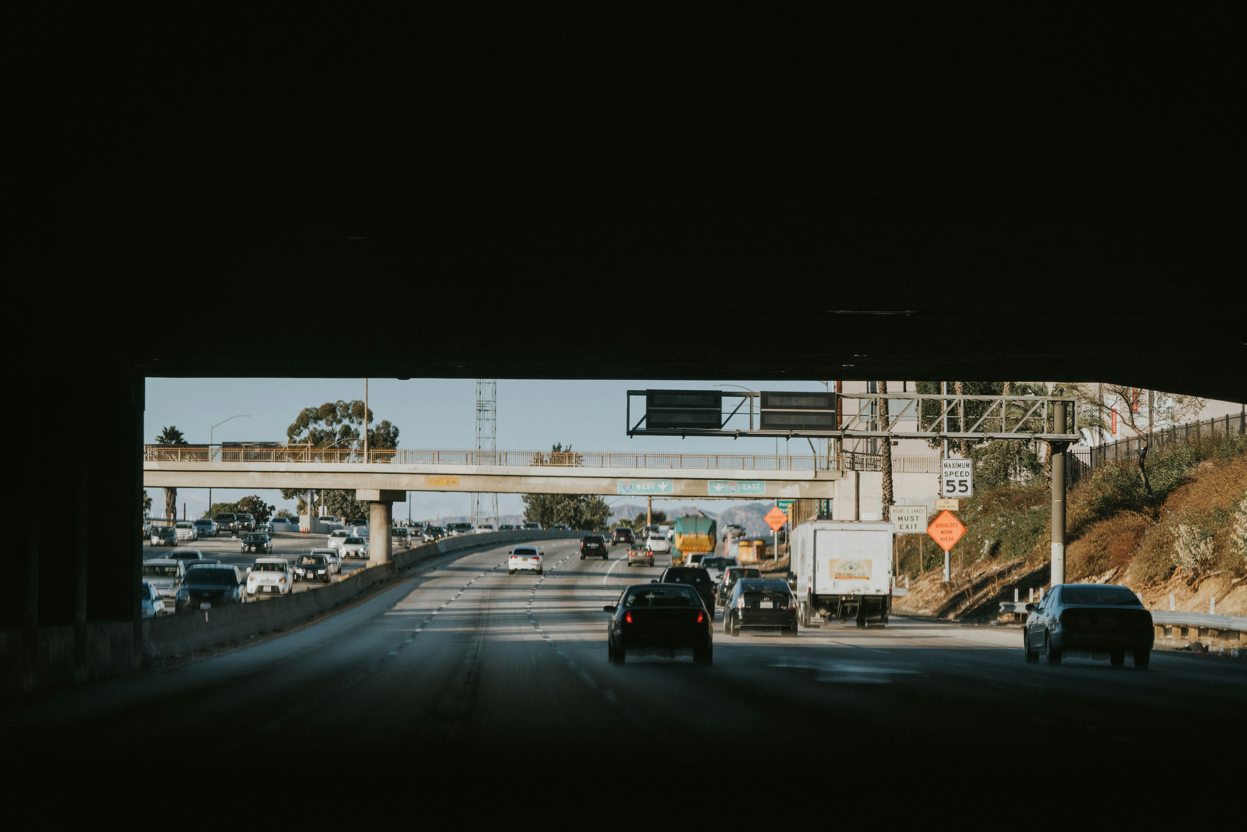 Traffic flows smoothly beneath a shadowy overpass, with glimpses of greenery and urban structures in the background.