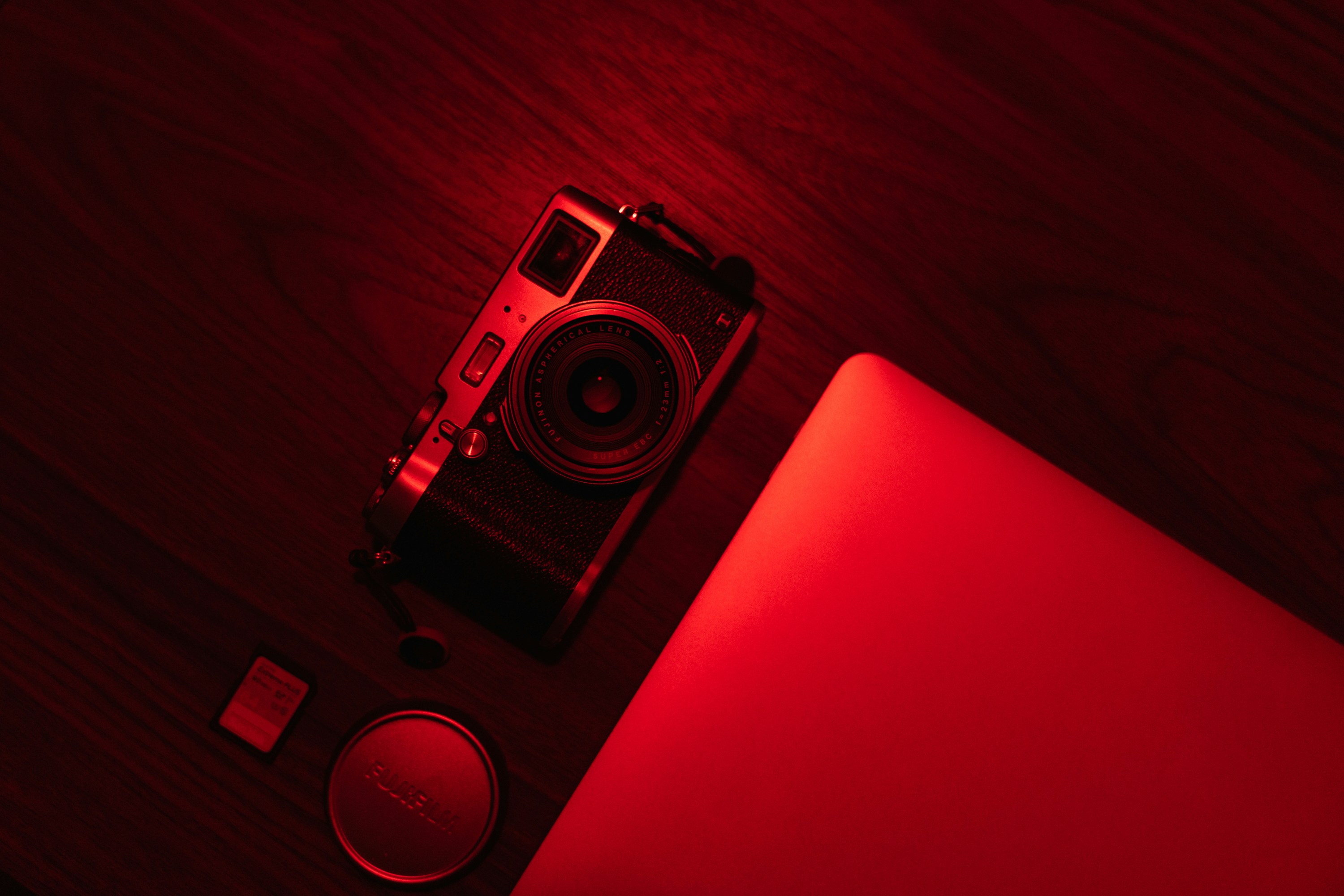 Vintage camera resting beside a laptop and memory card on a wooden surface illuminated by soft red light.