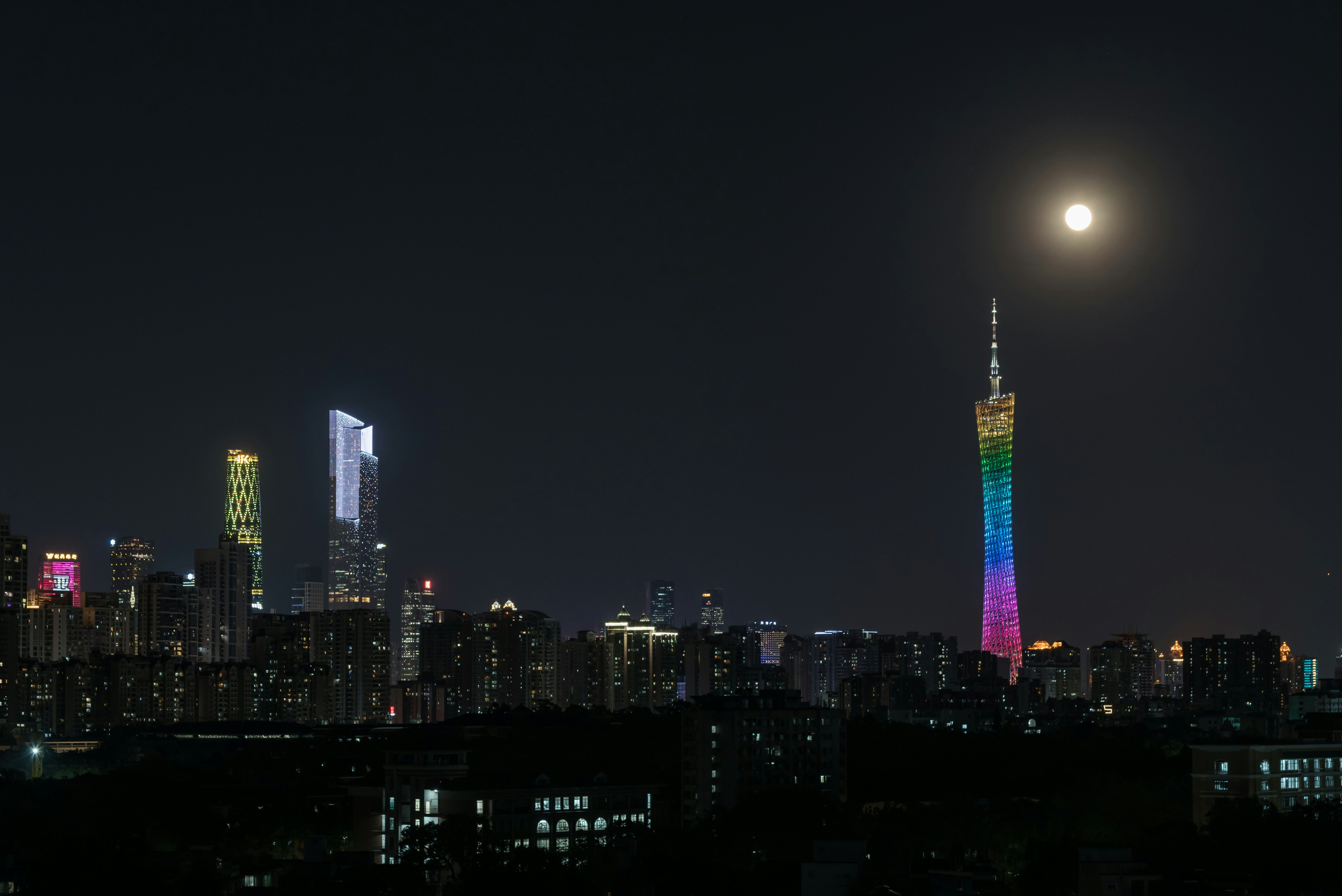 The Guangzhou Tower stands proudly illuminated against a night sky, with a full moon casting a soft glow over the vibrant cityscape.