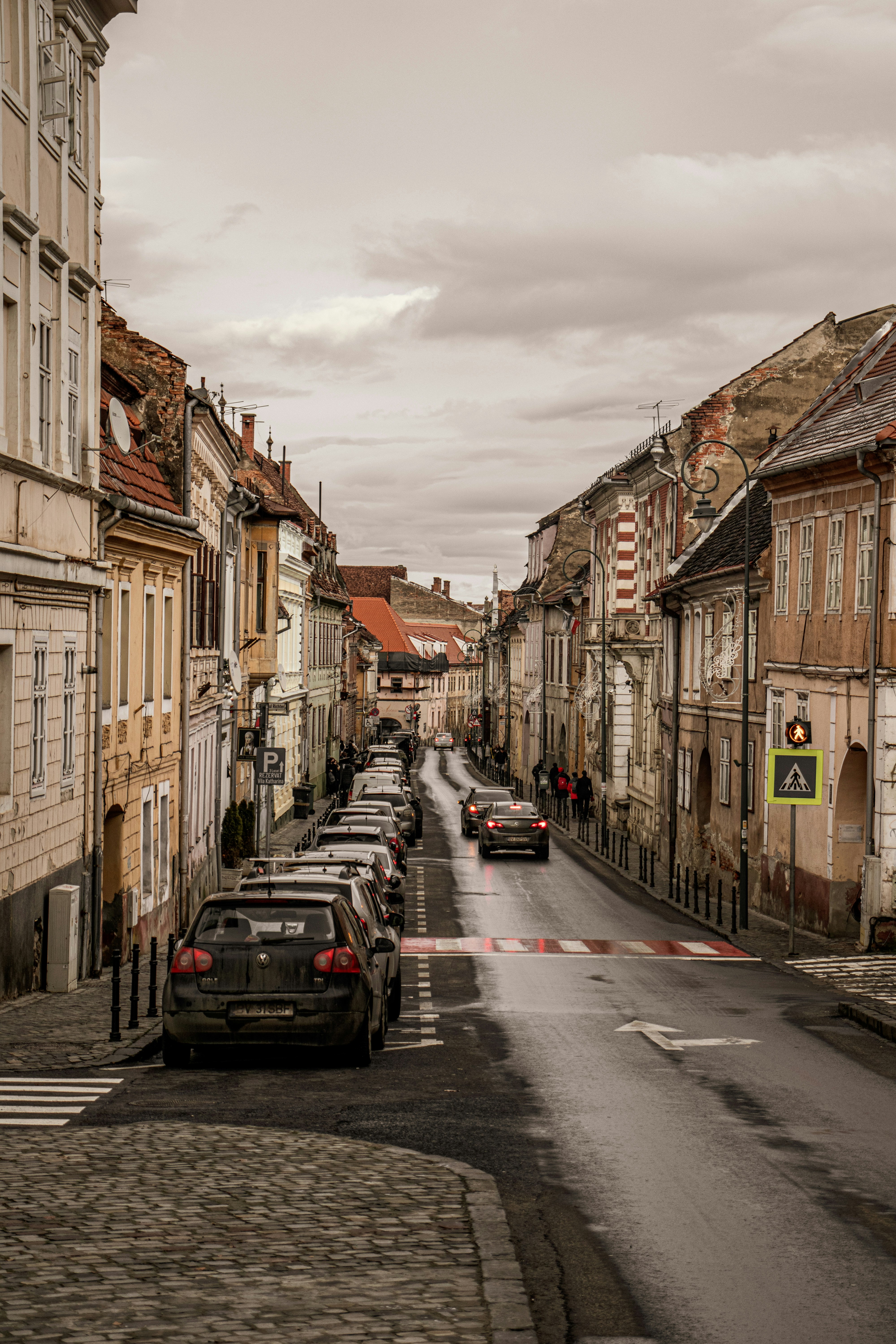 Narrow, cobblestone street lined with colorful historic buildings, showcasing parked cars and pedestrians under a cloudy sky.