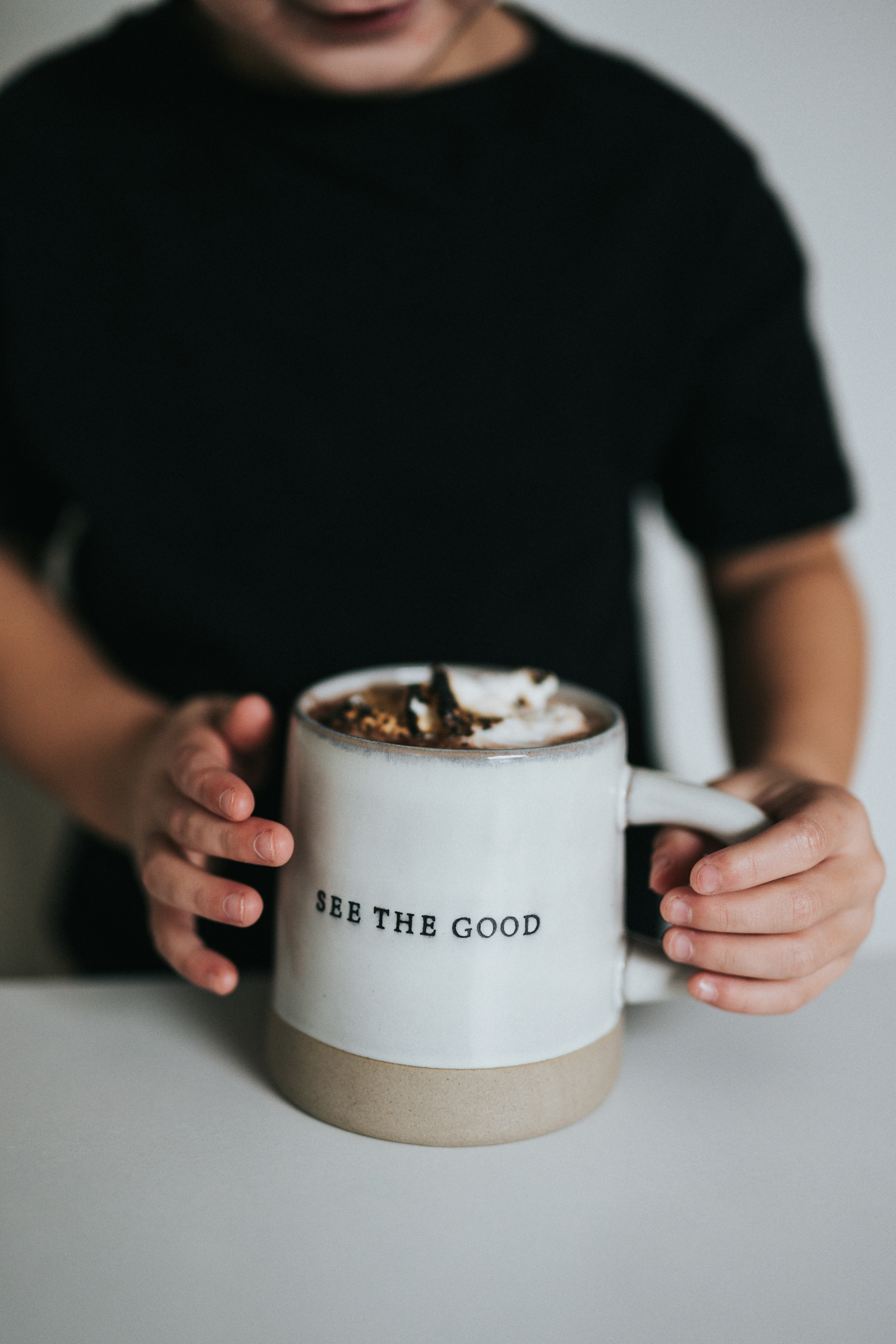 Child's hands gently holding a mug with the phrase 'SEE THE GOOD,' topped with whipped cream and chocolate sprinkles.