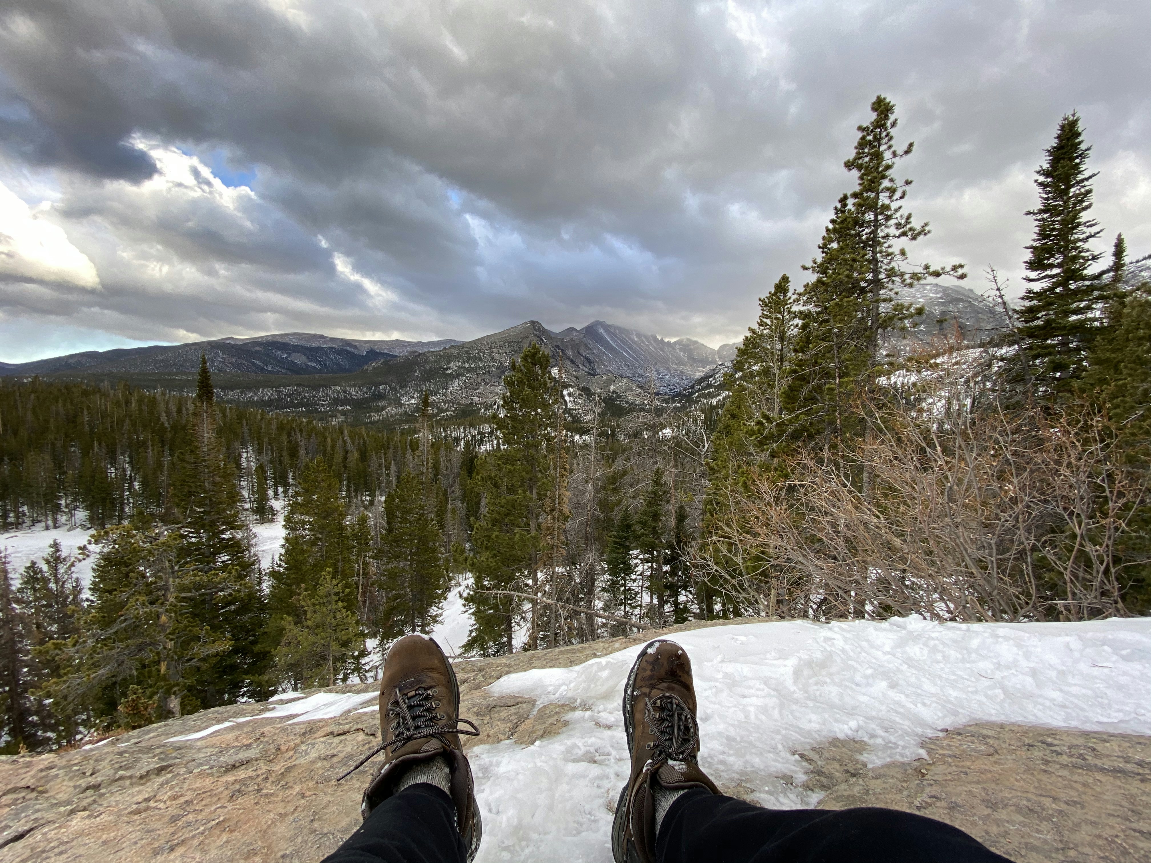 A pair of rugged boots resting on a rocky ledge, overlooking a vast snow-covered landscape dotted with evergreen trees and distant mountains under a dramatic sky.