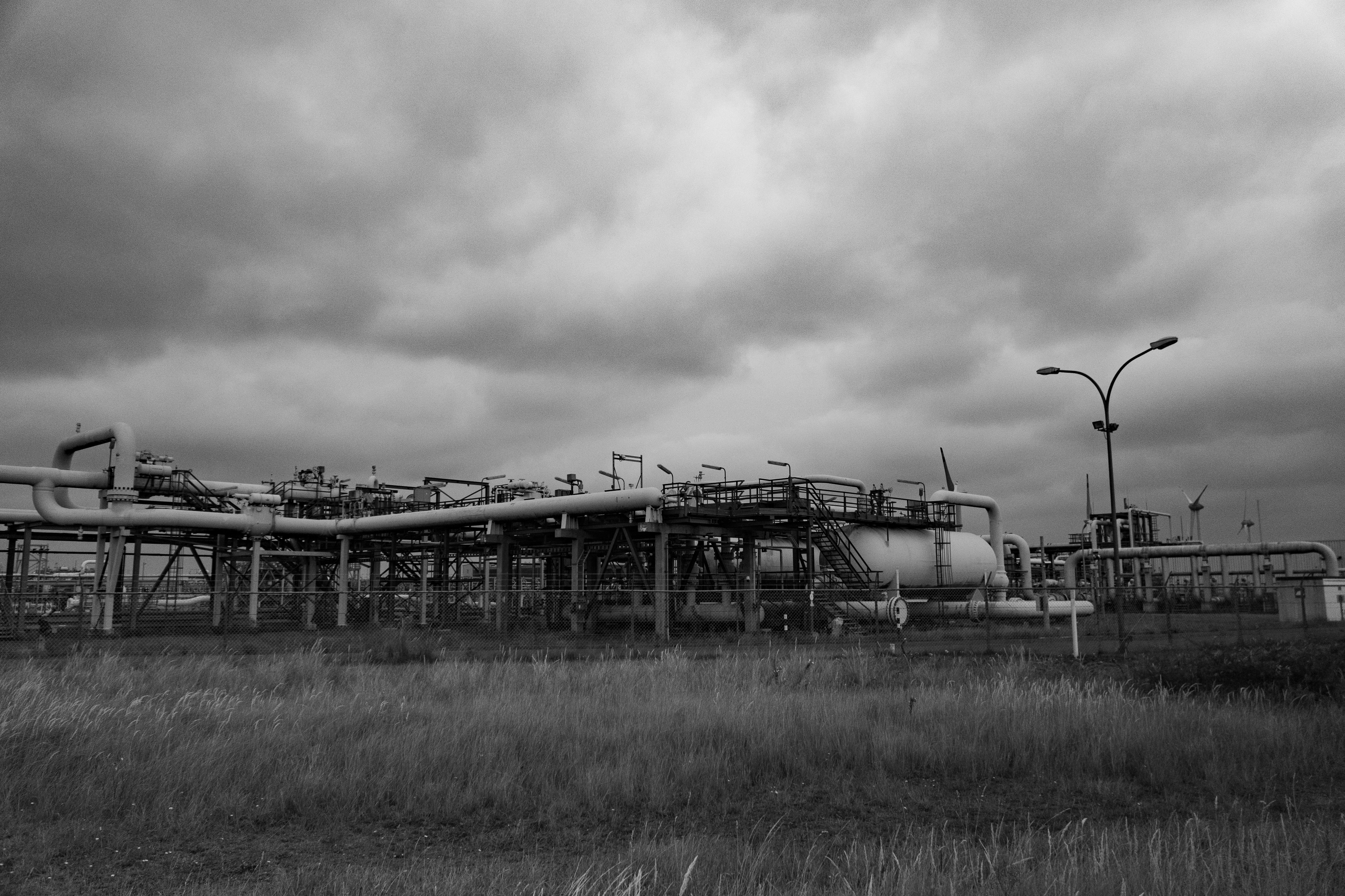 A complex network of industrial pipes and structures under a dramatic sky, showcasing the intricacies of modern engineering. The scene is rendered in monochrome, emphasizing texture and form.
