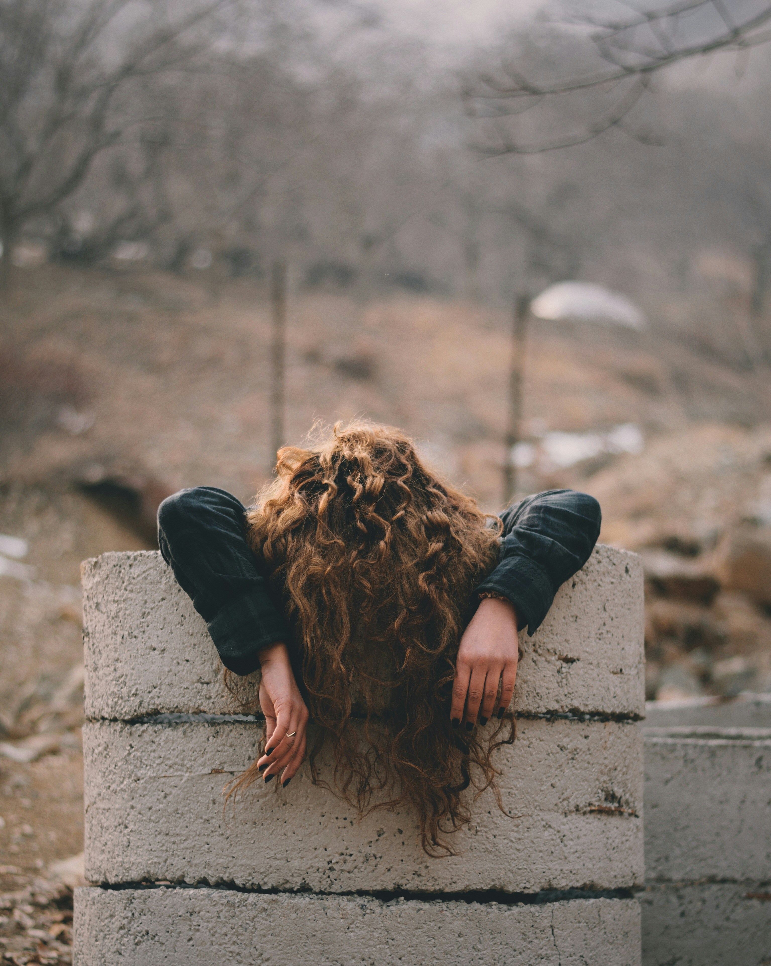 A woman with curly hair leans over a concrete barrier, surrounded by a muted landscape of trees and earth. Her relaxed posture evokes a sense of introspection.