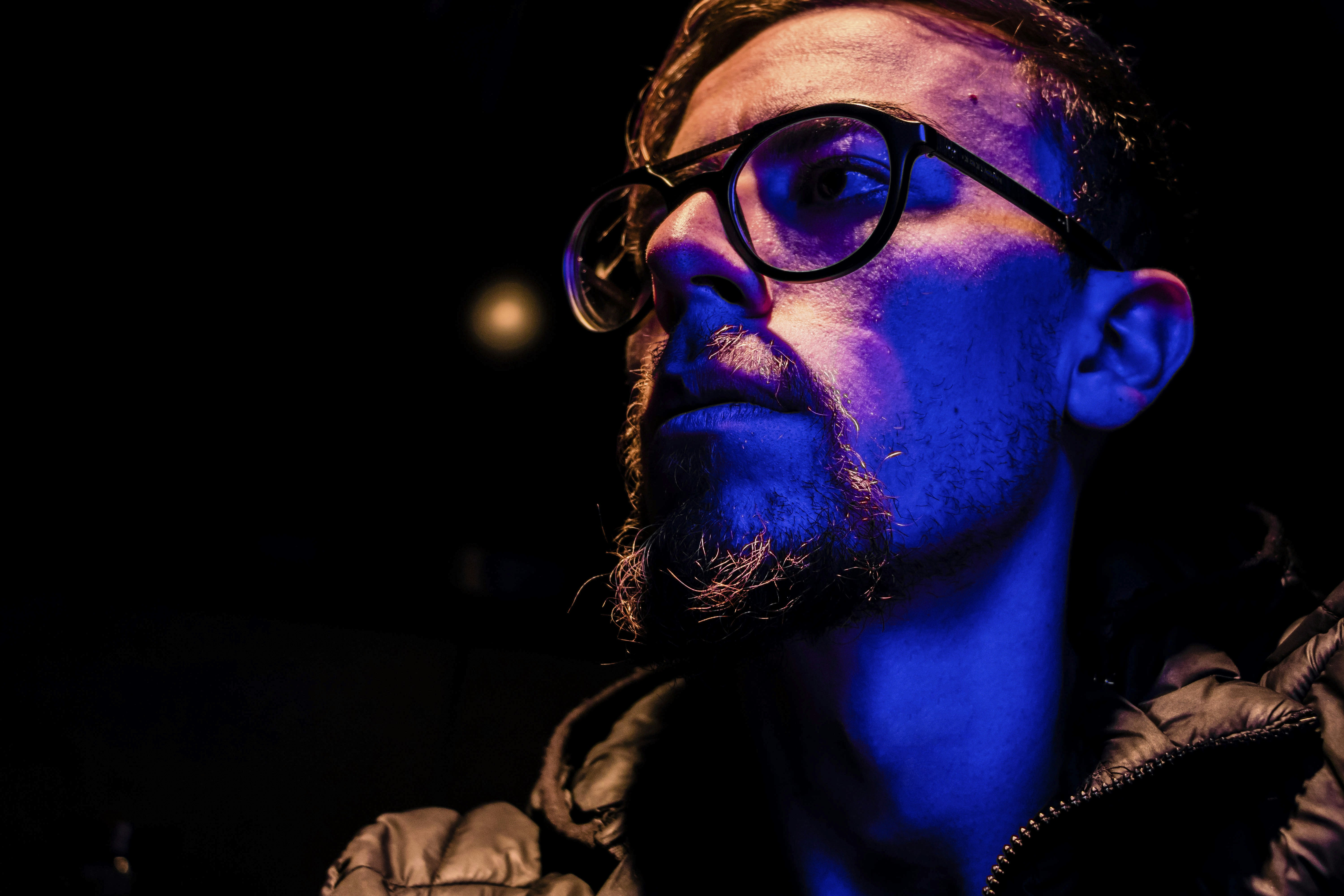 Man with glasses illuminated by blue and purple lights against a dark background.