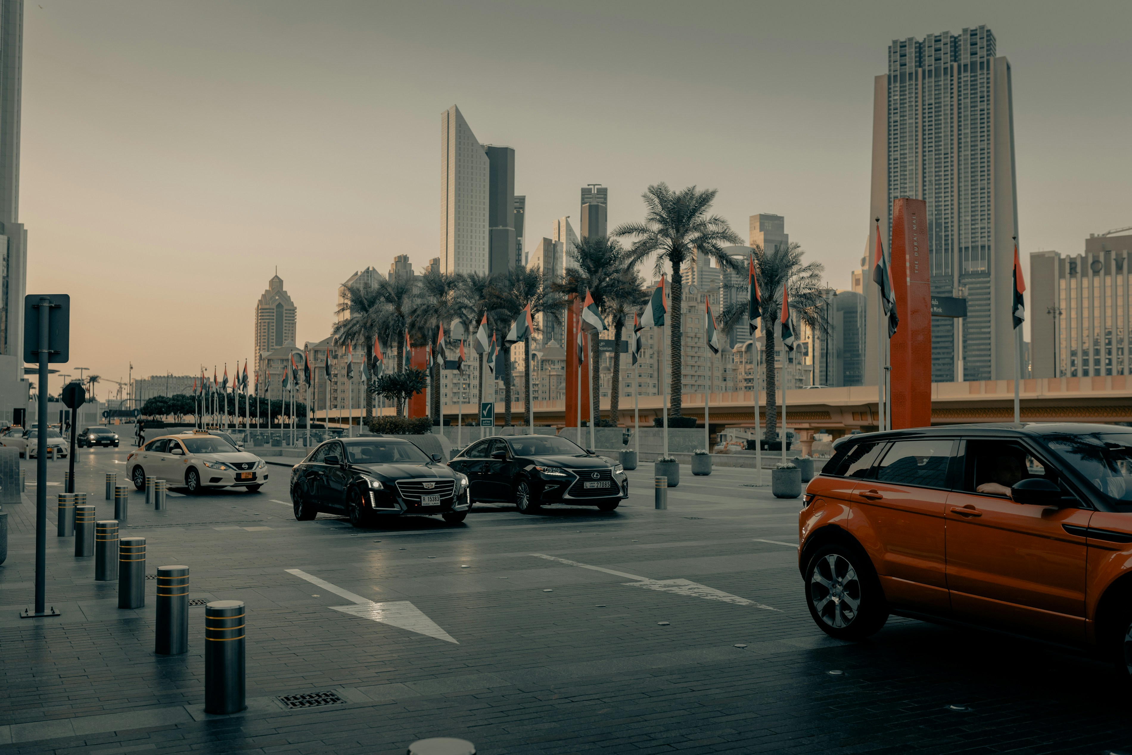 Cars navigate a bustling urban street lined with palm trees and flags, framed by modern skyscrapers in the background.